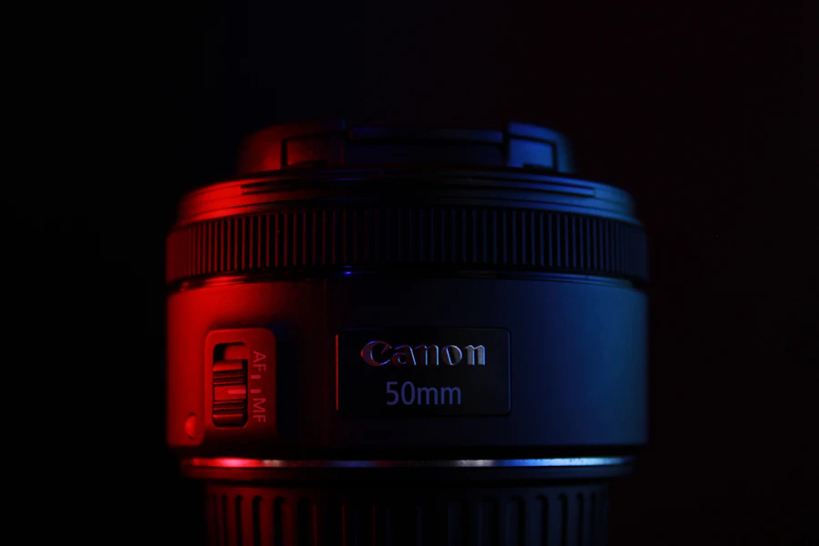 Canon 50mm lens photographed against a black background, lit dramatically with red light on the left and blue light on the right. The focus sits on the lens branding and barrel details.