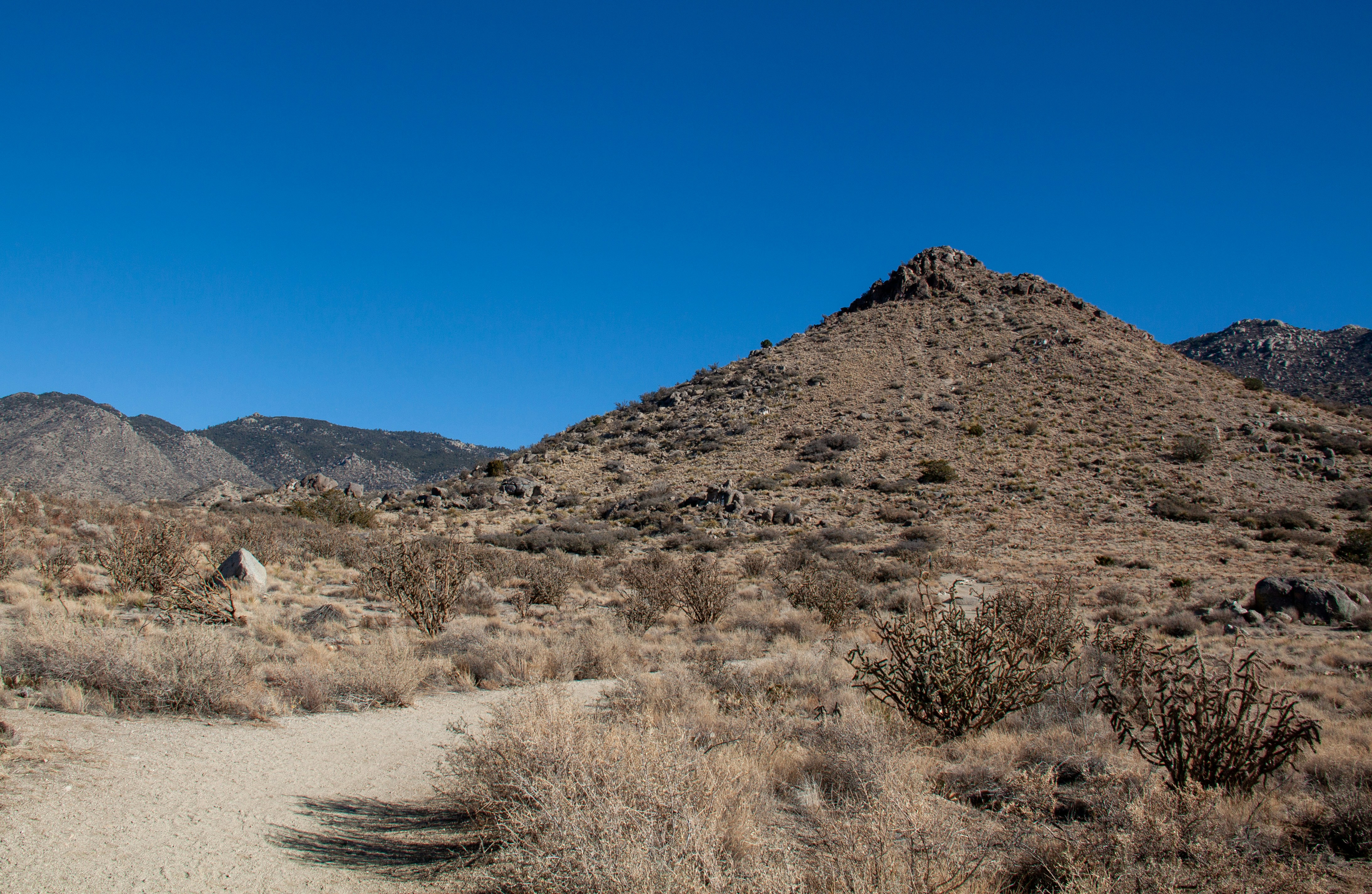 Arid mountain landscape with a sandy path winding through sparse vegetation beneath a deep blue sky.