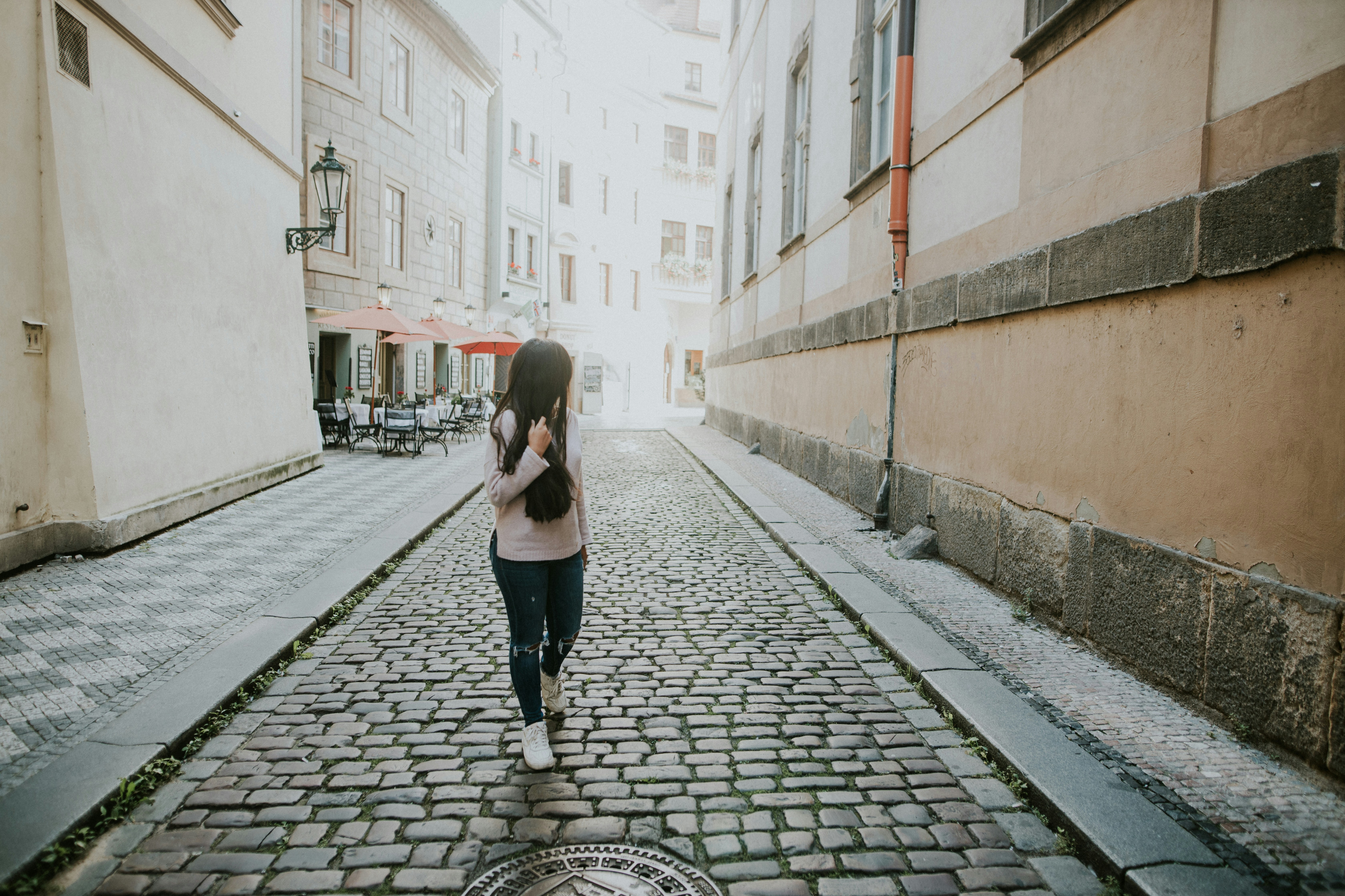 A young woman strolls down a quaint cobblestone street, flanked by historic buildings and outdoor seating. The scene captures a serene urban moment.