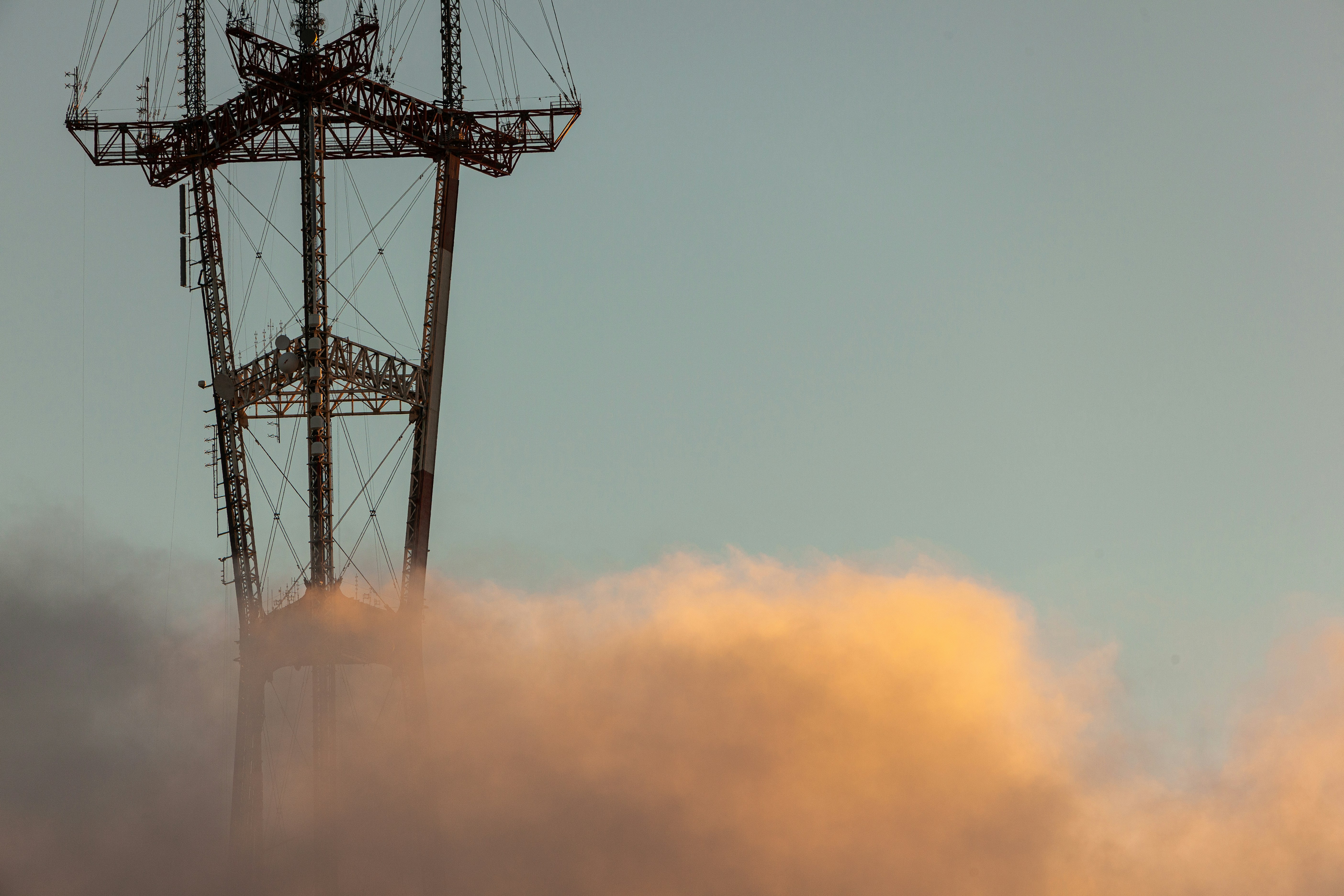 A towering communication structure rises above a blanket of soft, golden-hued clouds at dusk.