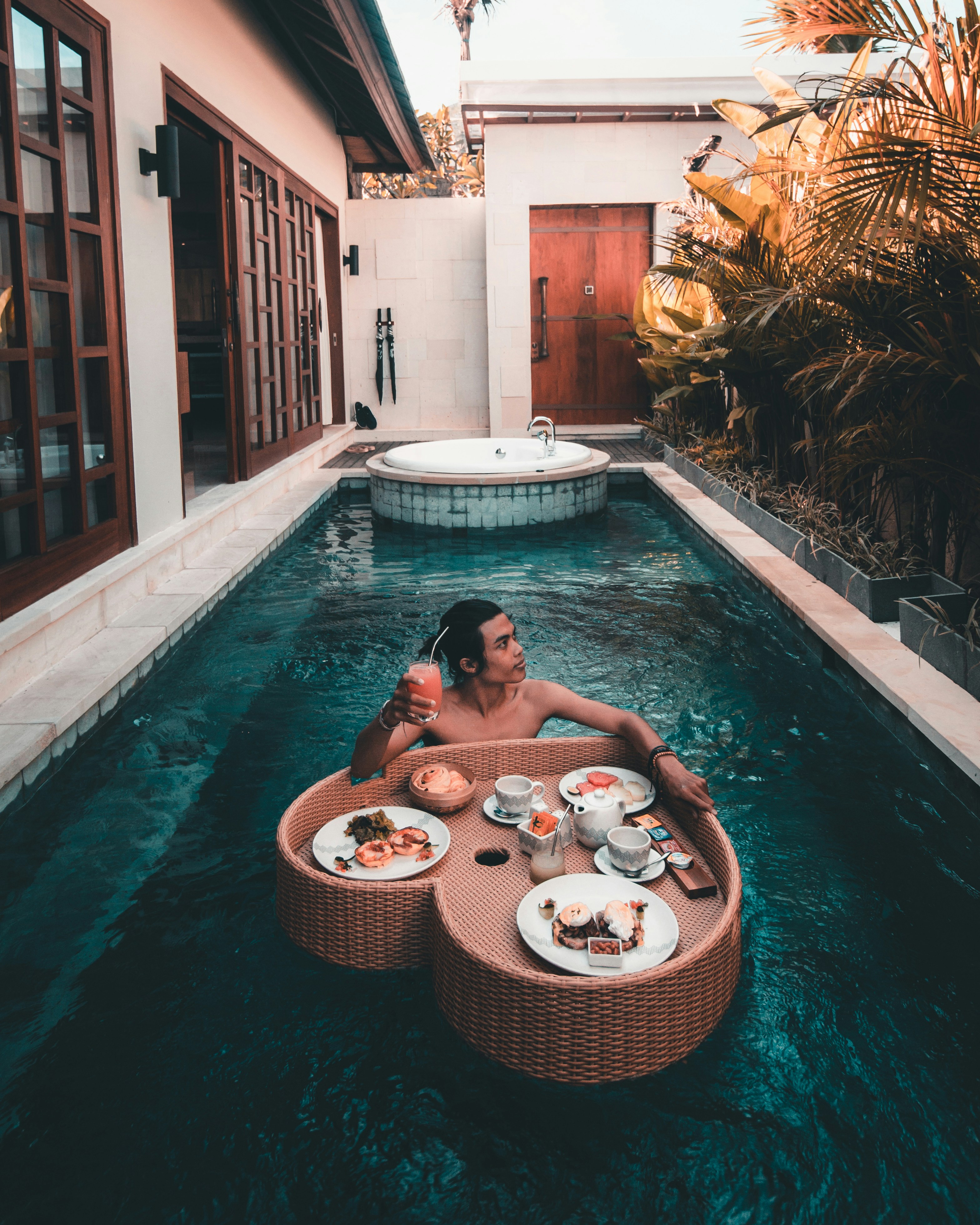 Man enjoying breakfast in a floating tray while relaxing in a tranquil pool surrounded by lush greenery and modern architecture.