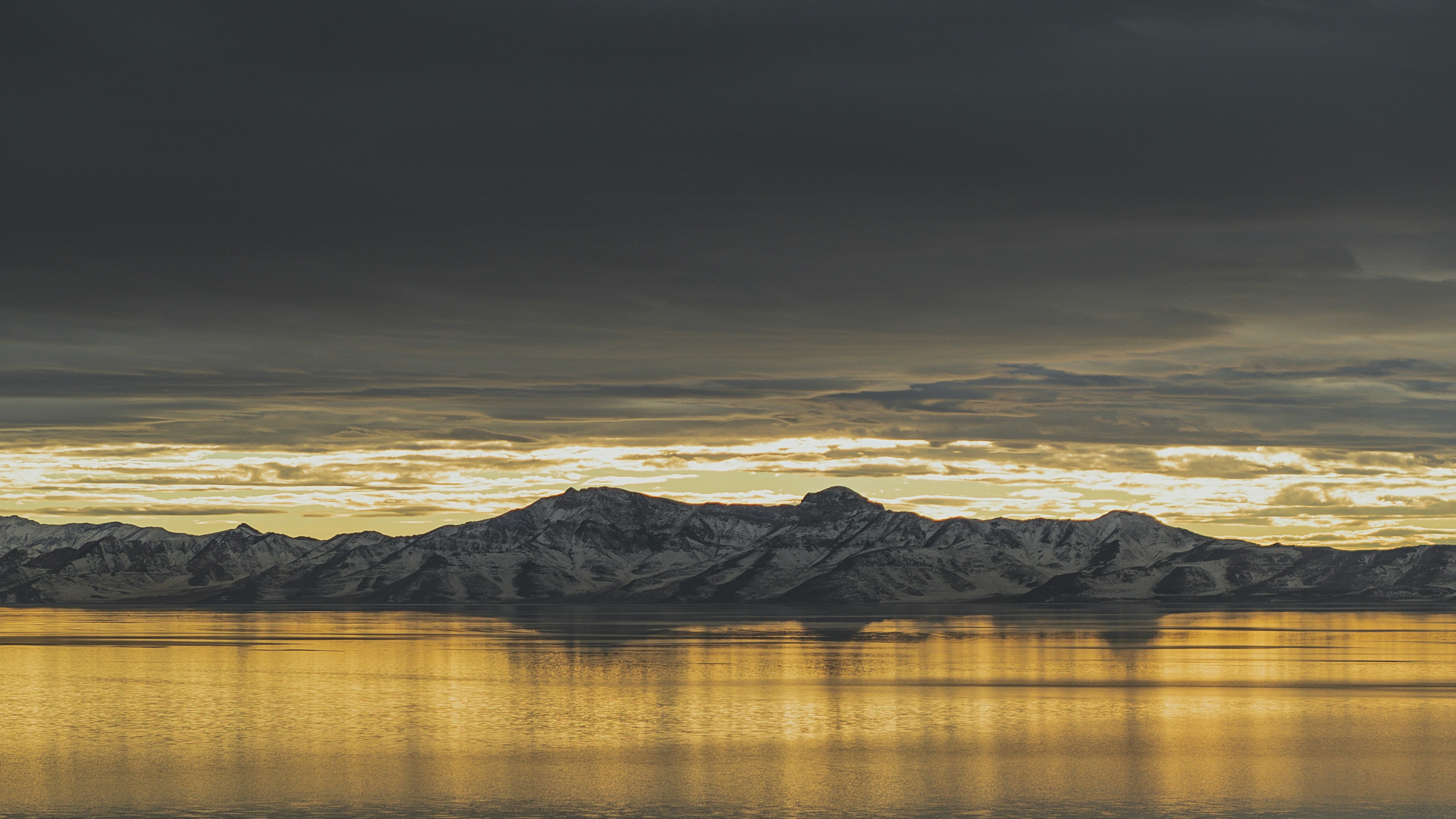 Golden reflections dance on a tranquil lake, framed by majestic mountains under a moody sky. The scene captures the serene transition of day to night.