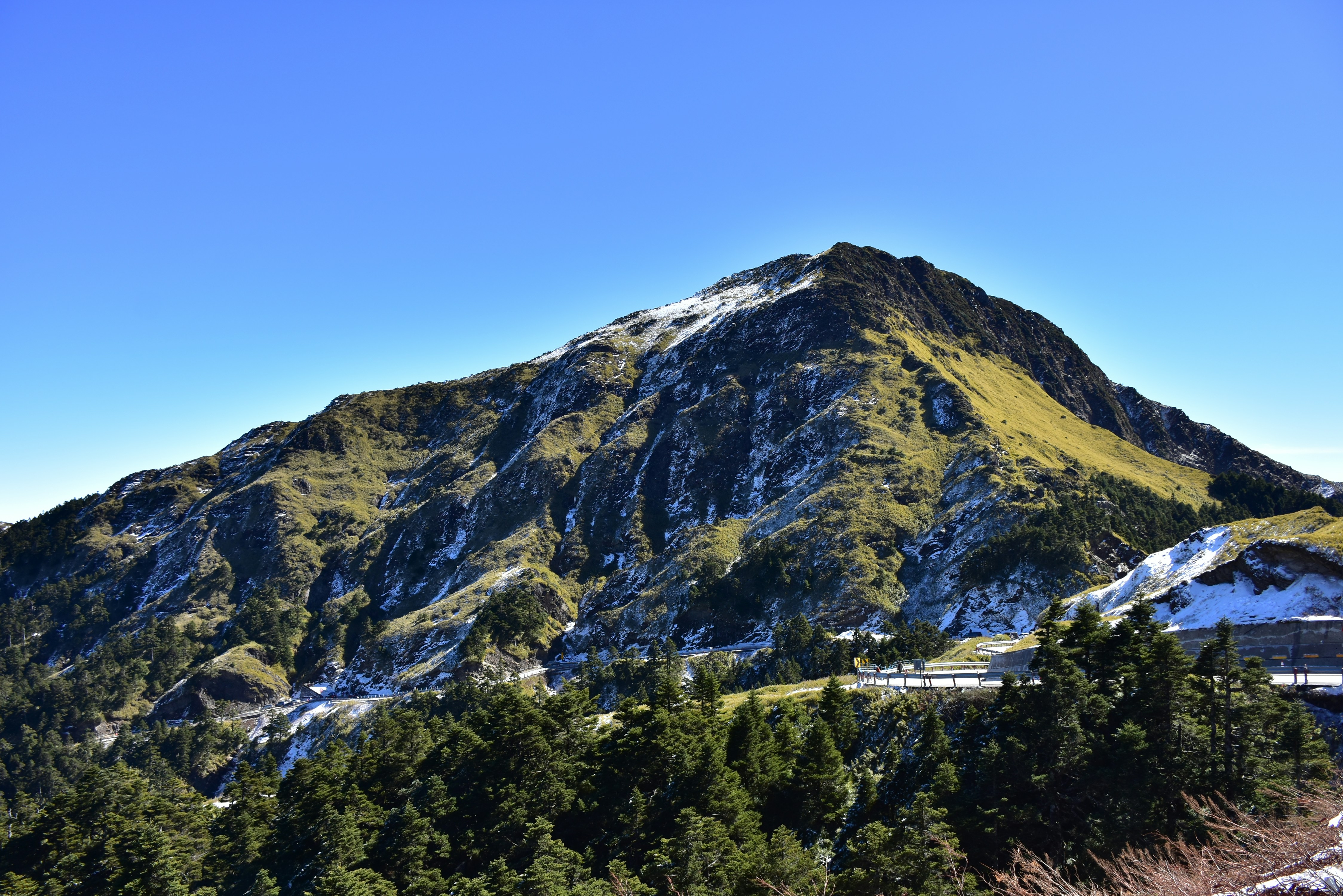 Lush green slopes meet rugged peaks under a clear blue sky, showcasing the natural beauty of mountainous terrain. Snow caps the higher elevations, adding a touch of winter's embrace.
