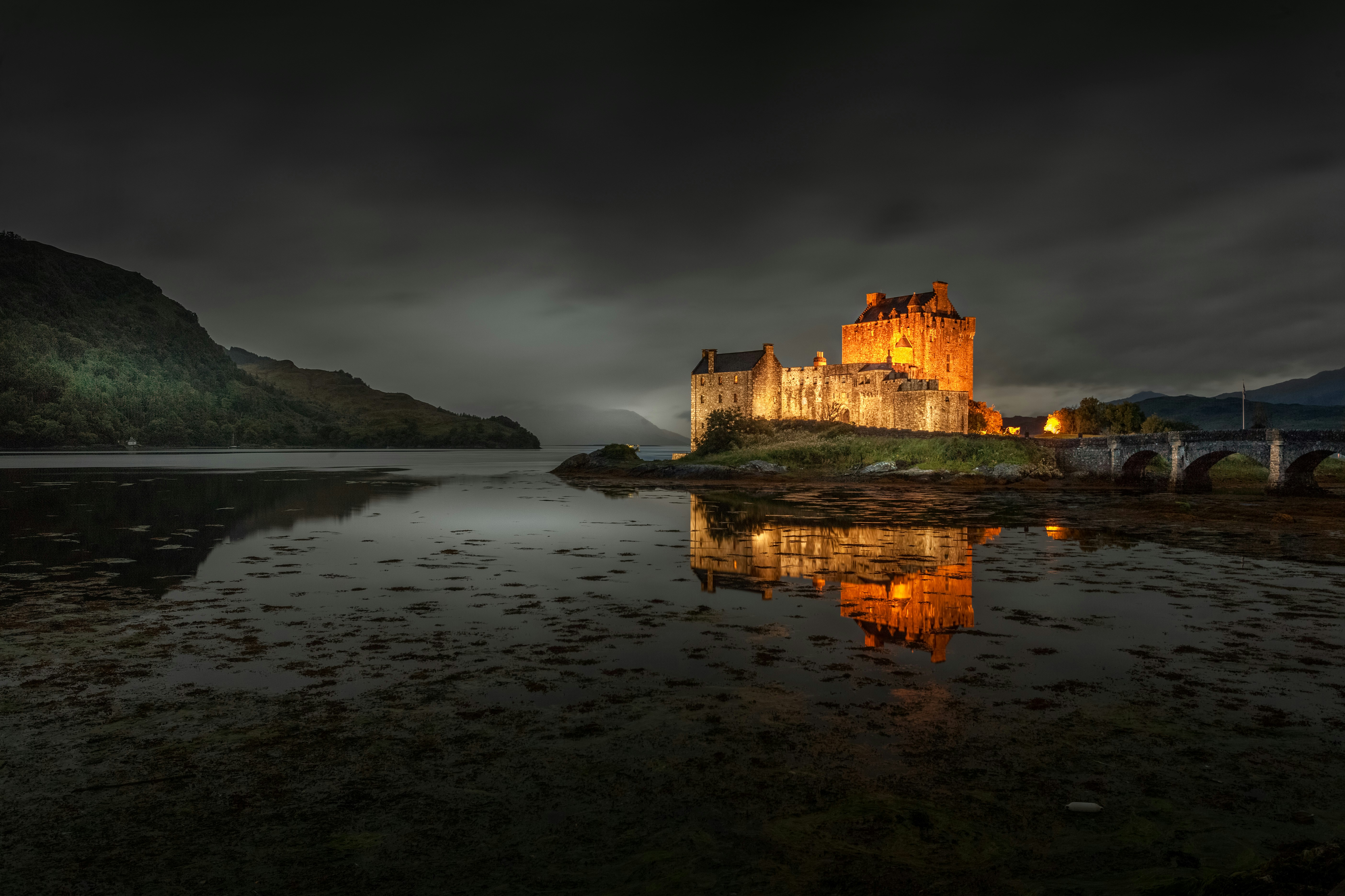 Historic castle reflecting in calm waters under a moody sky, illuminated by warm lights. The scene captures the essence of ancient architecture amidst natural beauty.