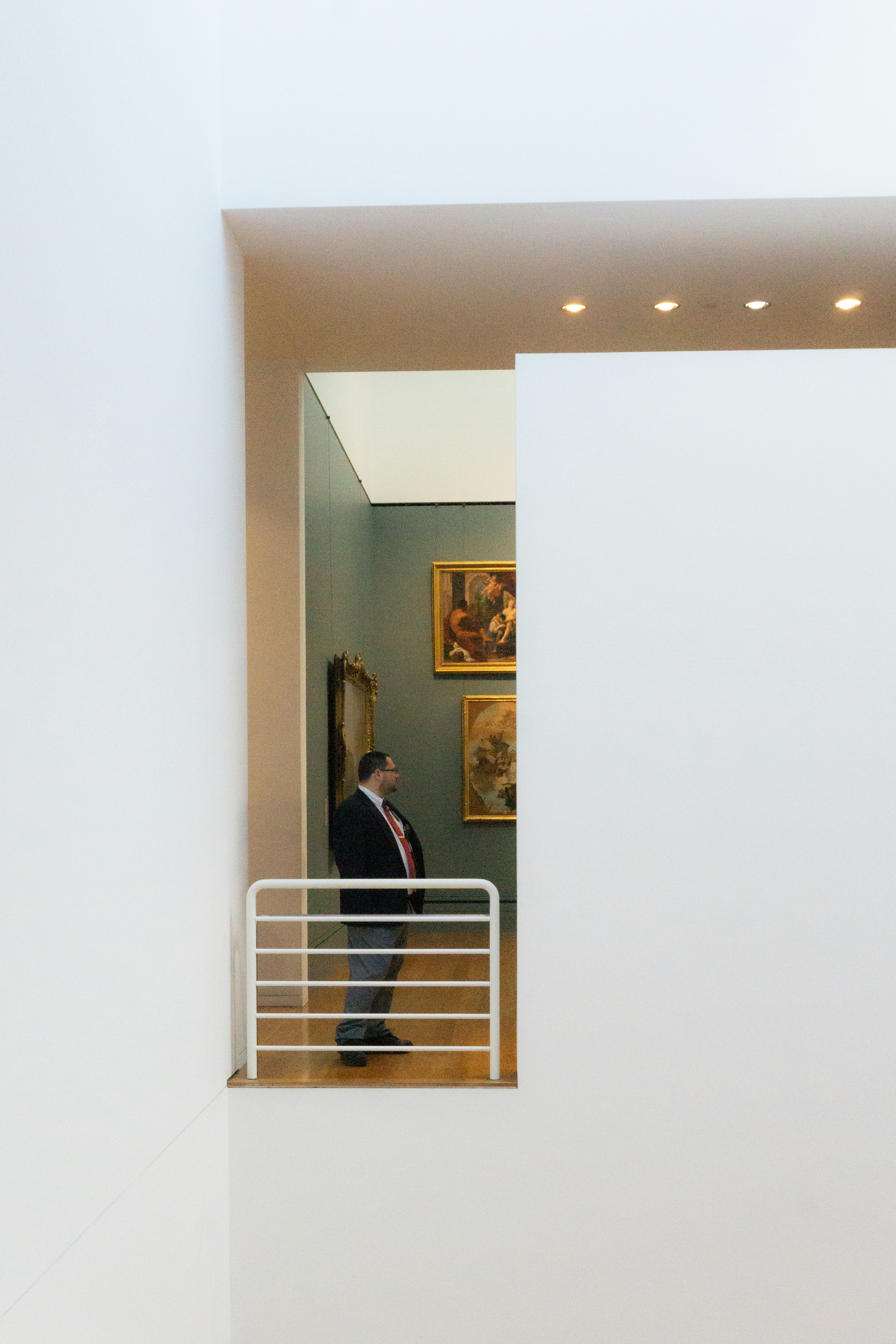 A man stands quietly in a modern art gallery, framed by minimalist architecture and classic paintings. The contrast between the contemporary space and the historical artworks creates a unique visual dialogue.