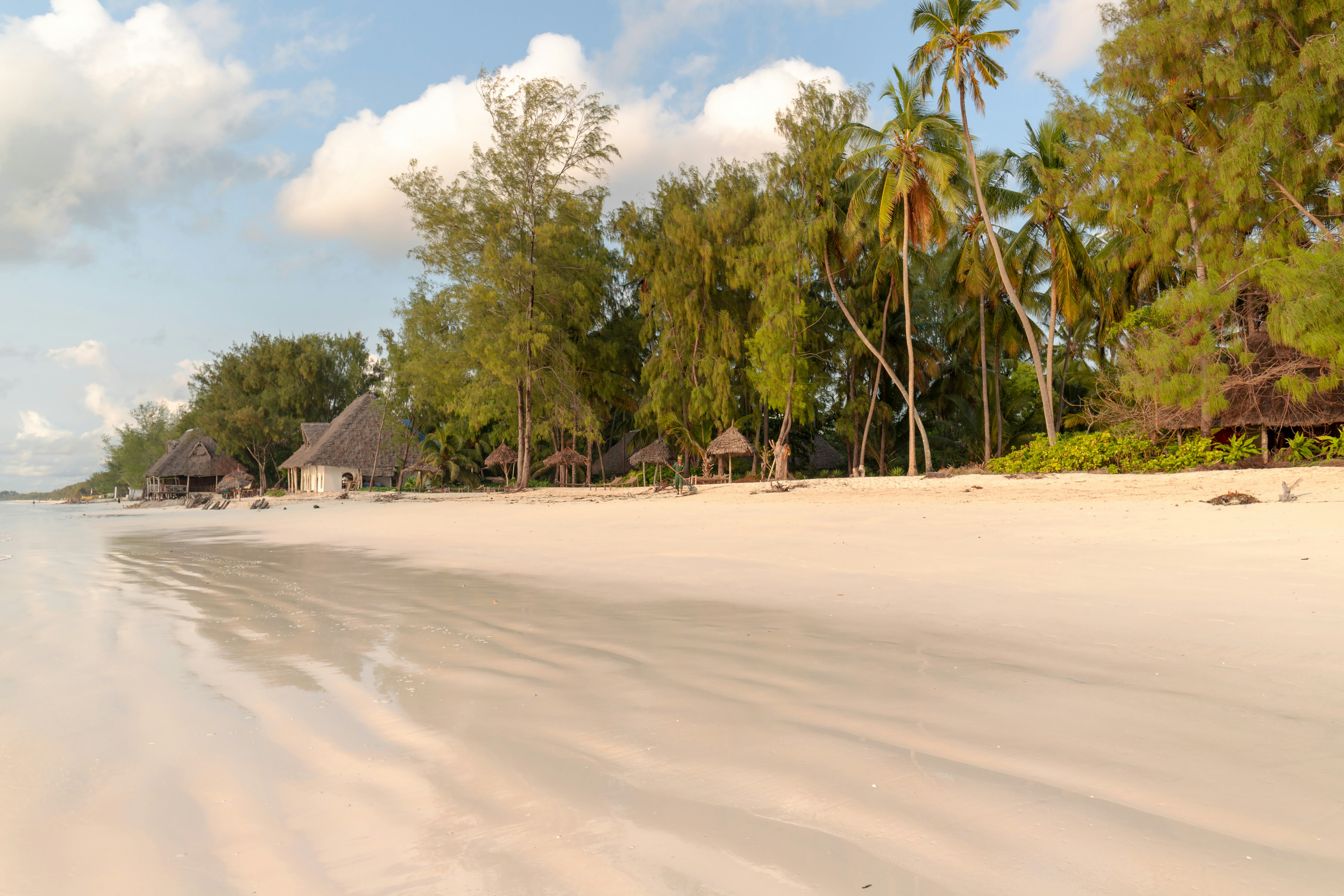 Serene beach with gentle waves, lined with lush trees and thatched huts under a clear sky.