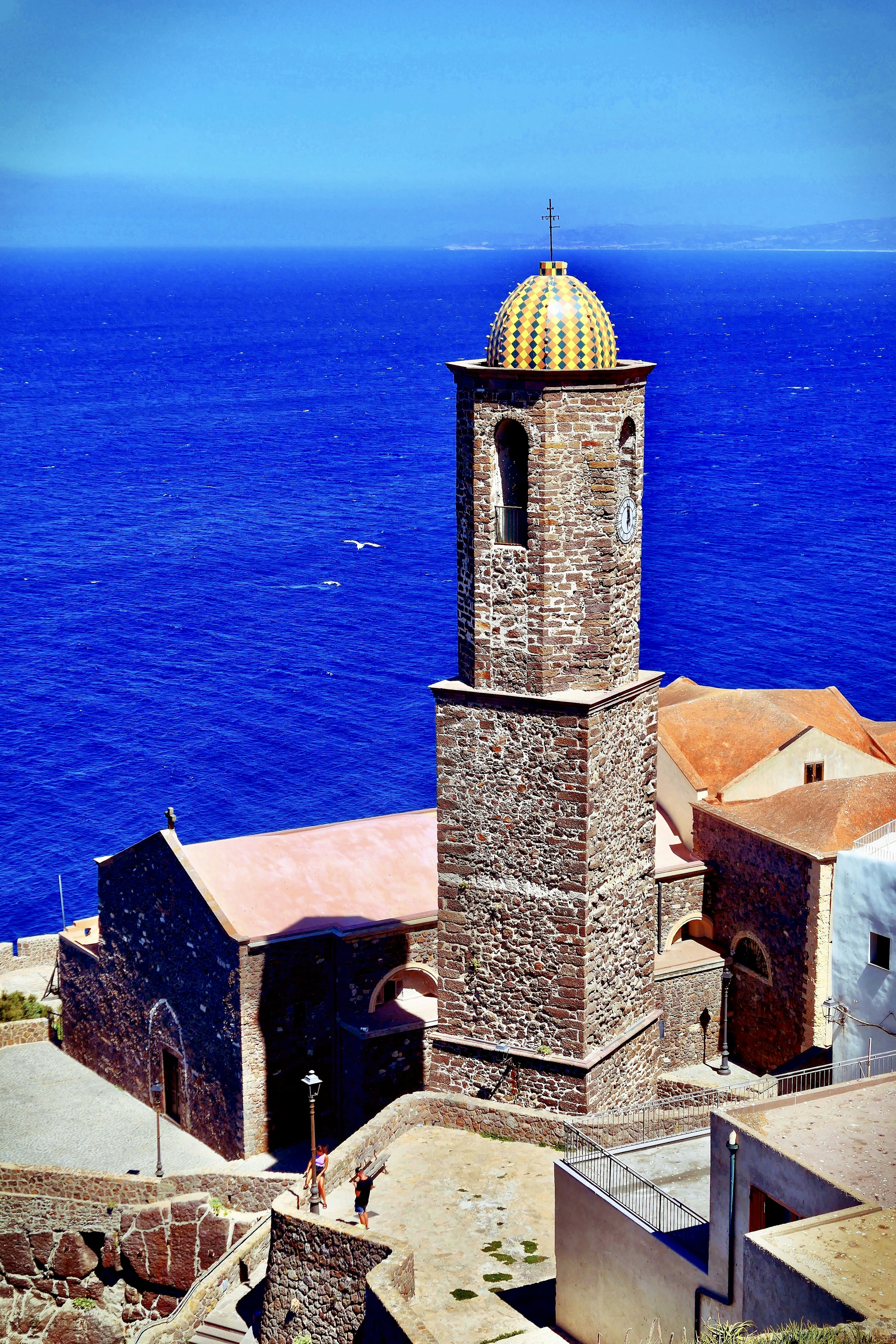 Historic clock tower with a golden dome stands against a vibrant blue sea, surrounded by charming architecture.