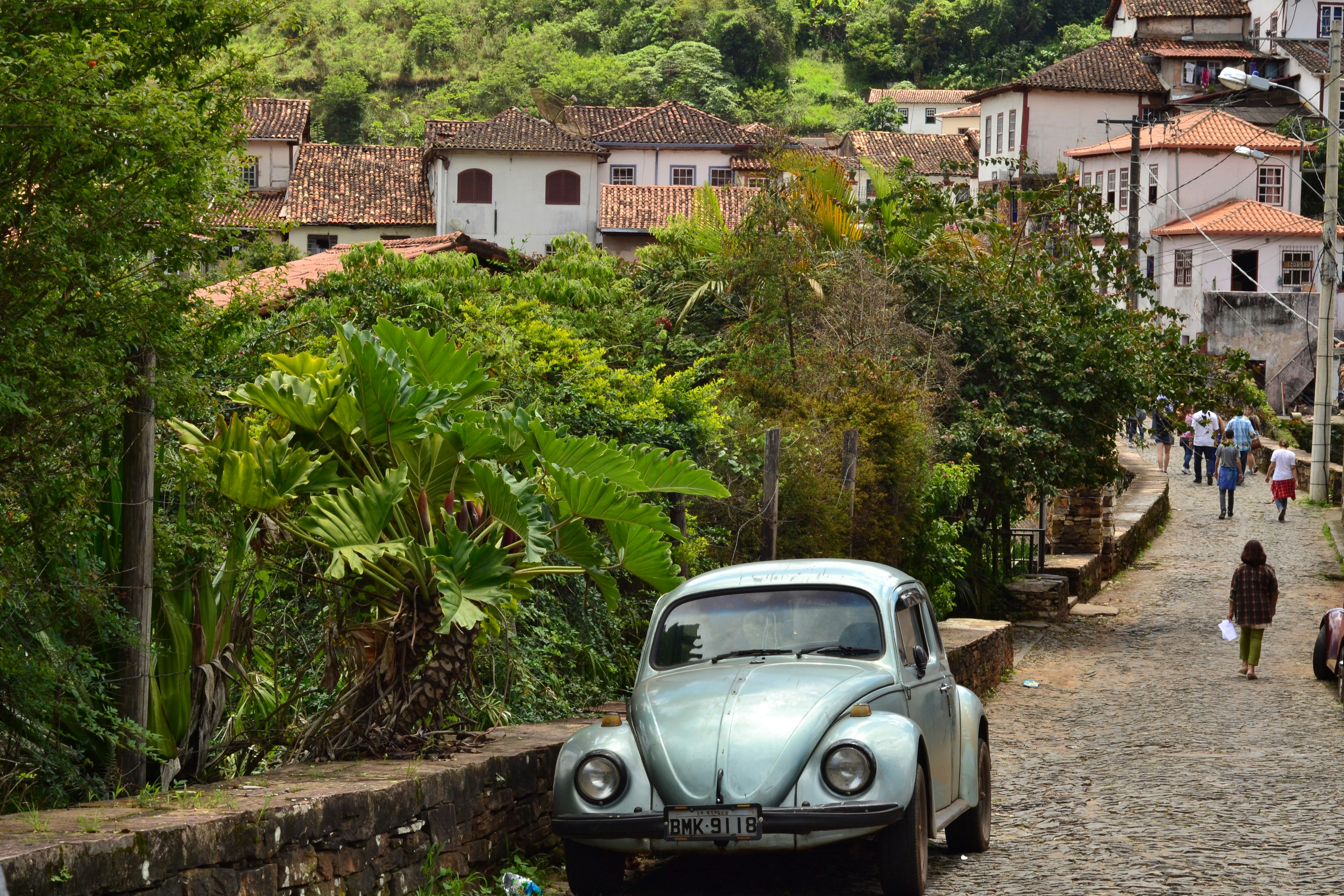 Green Volkswagen Beetle parked beside a lush, cobblestone street with hillside houses in the background.