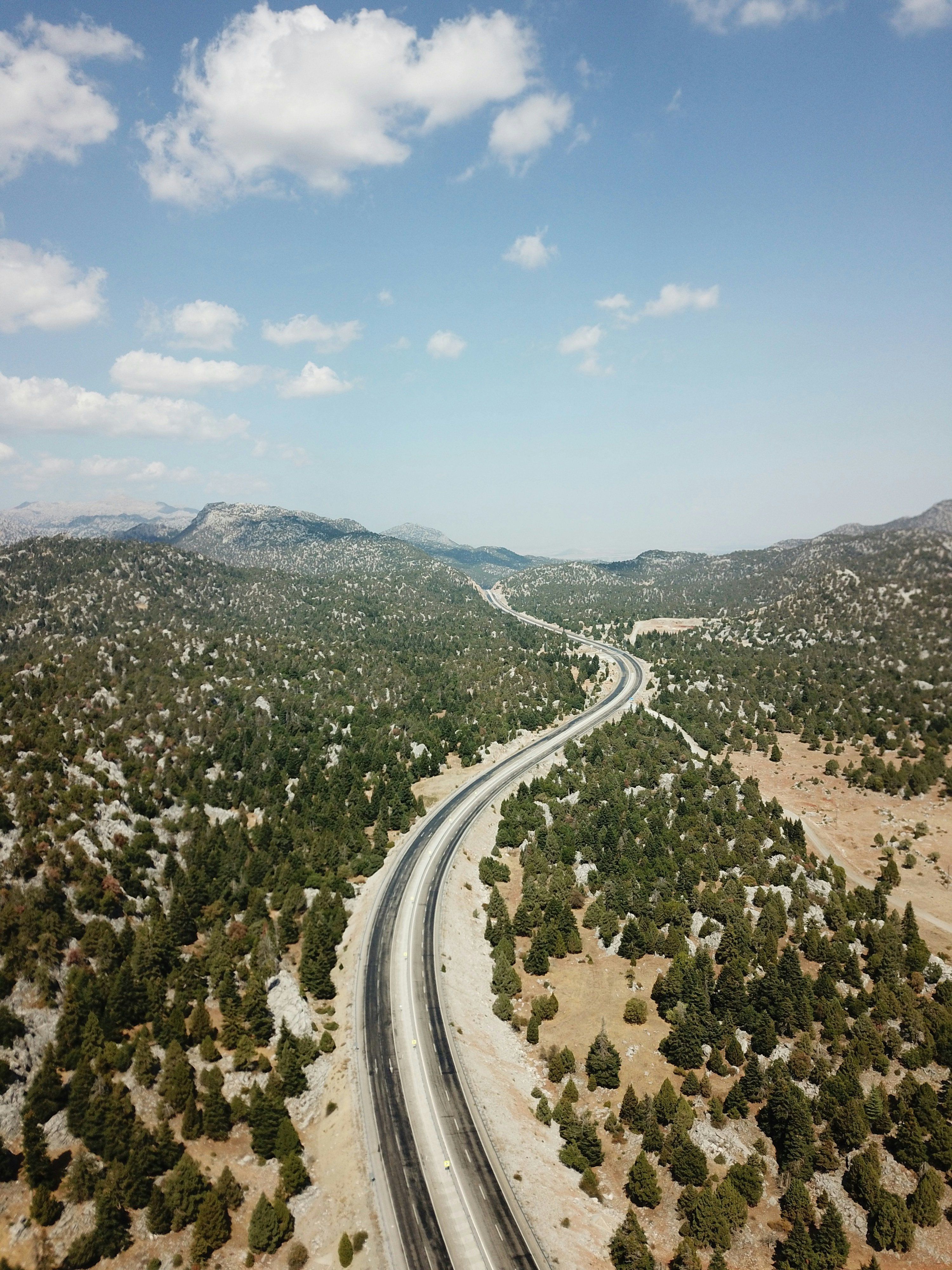 Aerial view of a winding highway cutting through a dense forest landscape, framed by mountains and a clear blue sky.