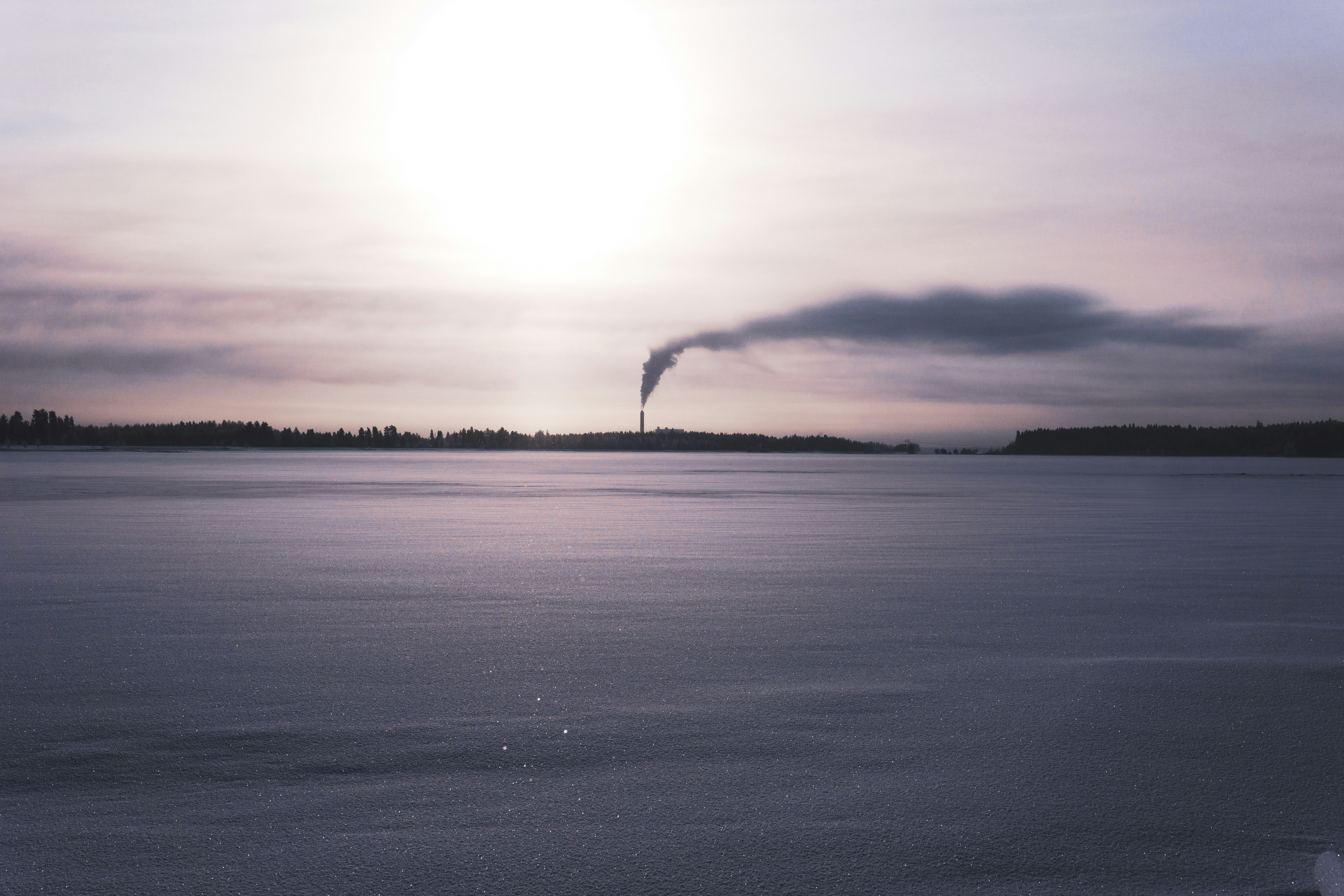 A tranquil winter landscape featuring a frozen lake under a soft sunset, with a distant smokestack releasing a plume of smoke into the sky.