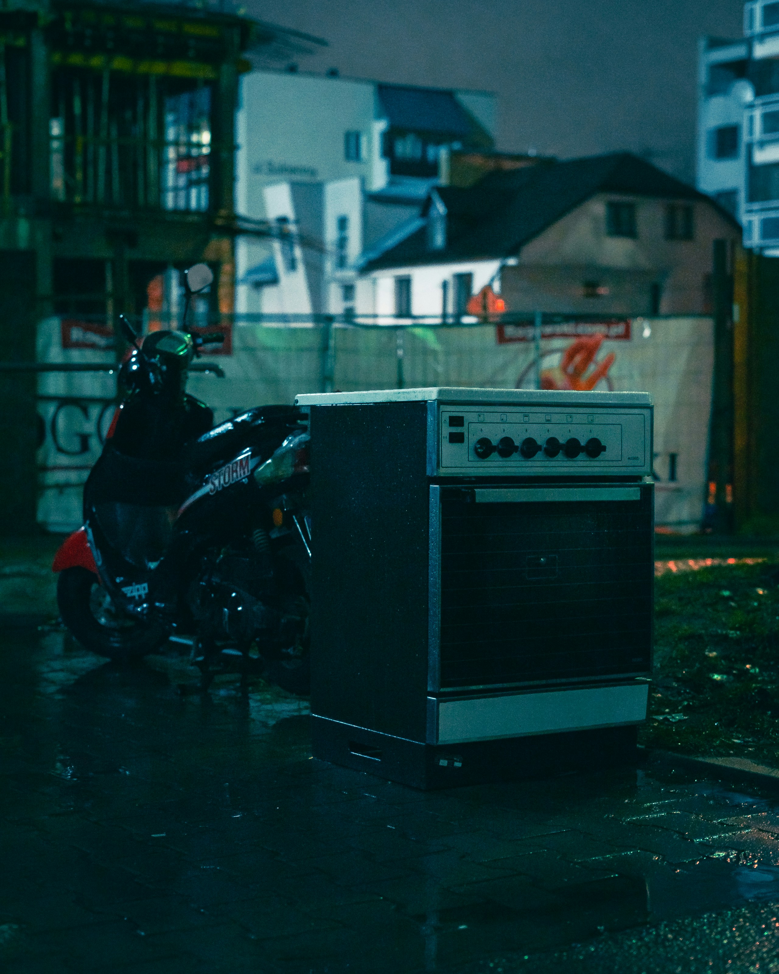Urban relic on rain-slick street: a vintage oven guards the motorcycle at midnight