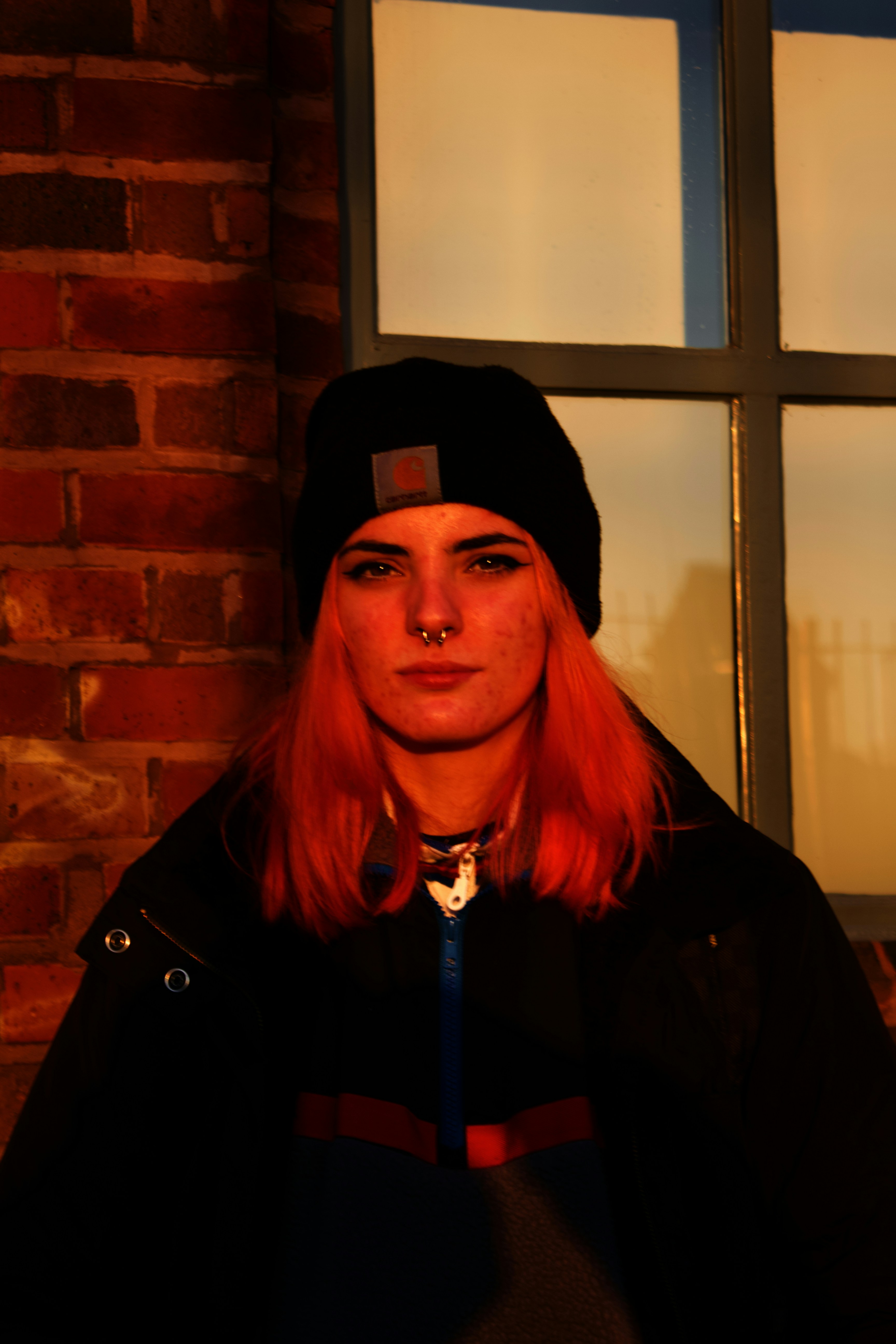 A young person with vibrant orange hair stands confidently in front of a brick wall, illuminated by warm sunlight. The scene captures a moment of urban expression.