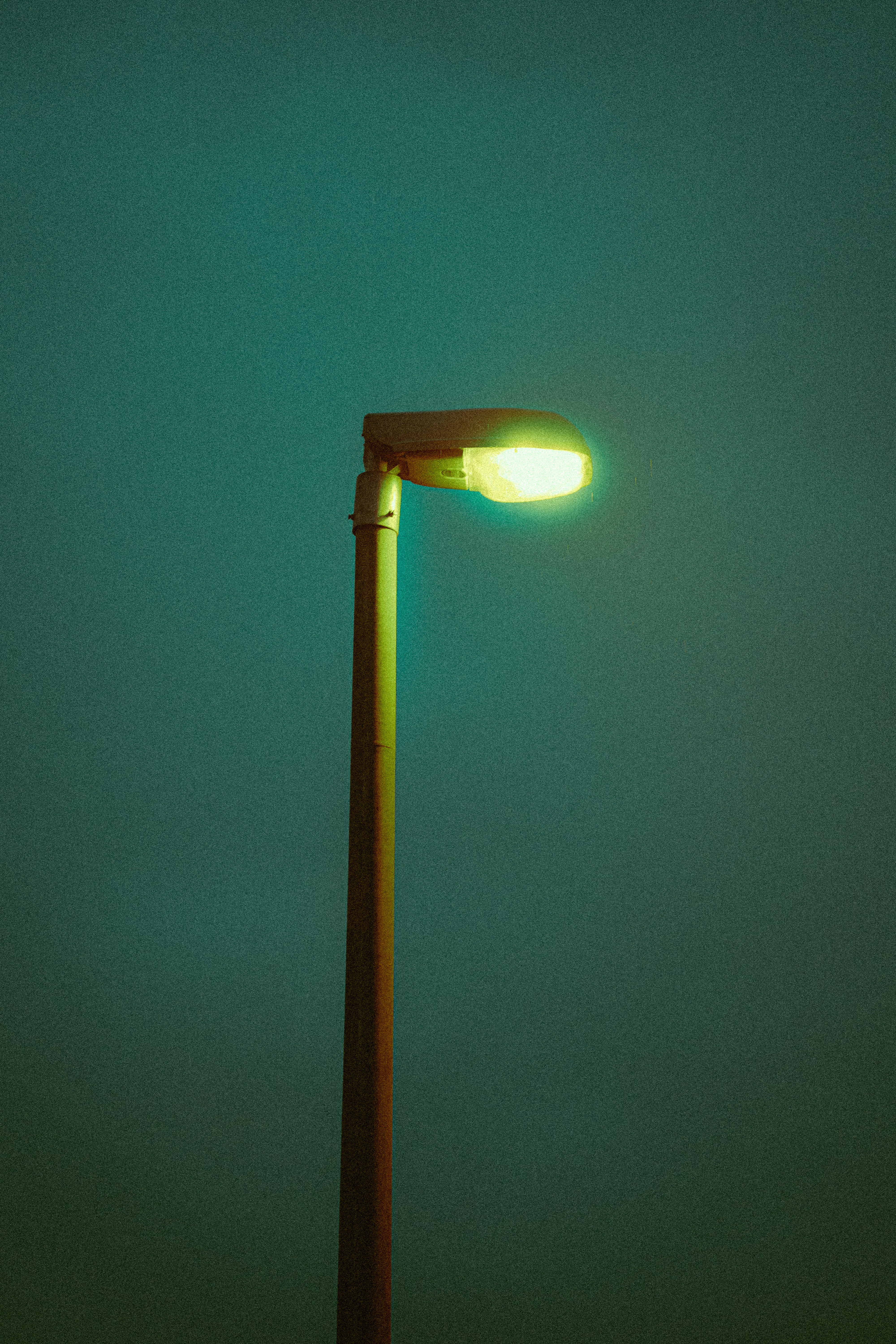 A solitary streetlight casts a soft green glow against a muted teal sky, highlighting its industrial design and creating a serene atmosphere.