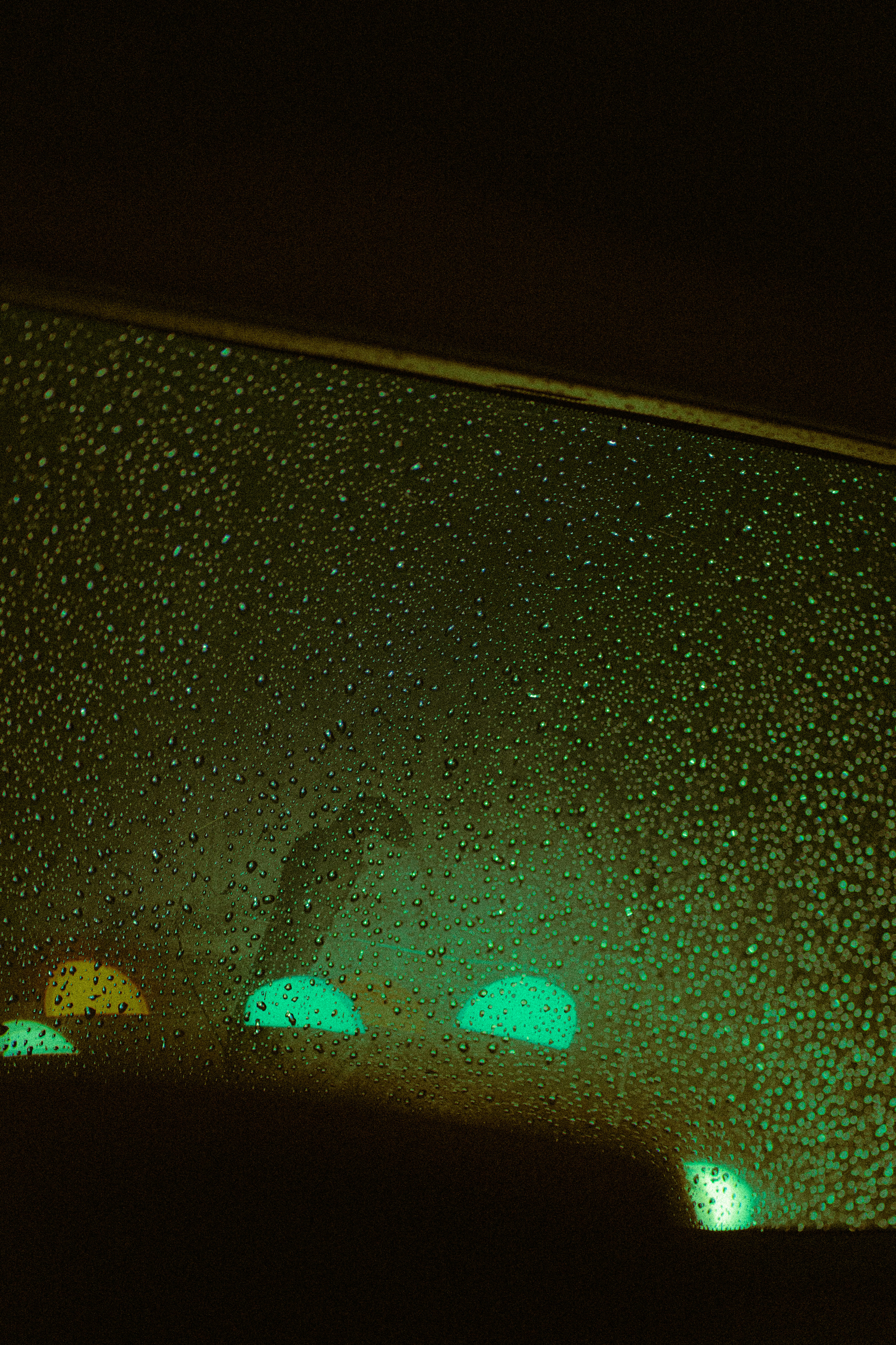 Raindrops scattered across a glass pane with blurred green and yellow lights in the background.
