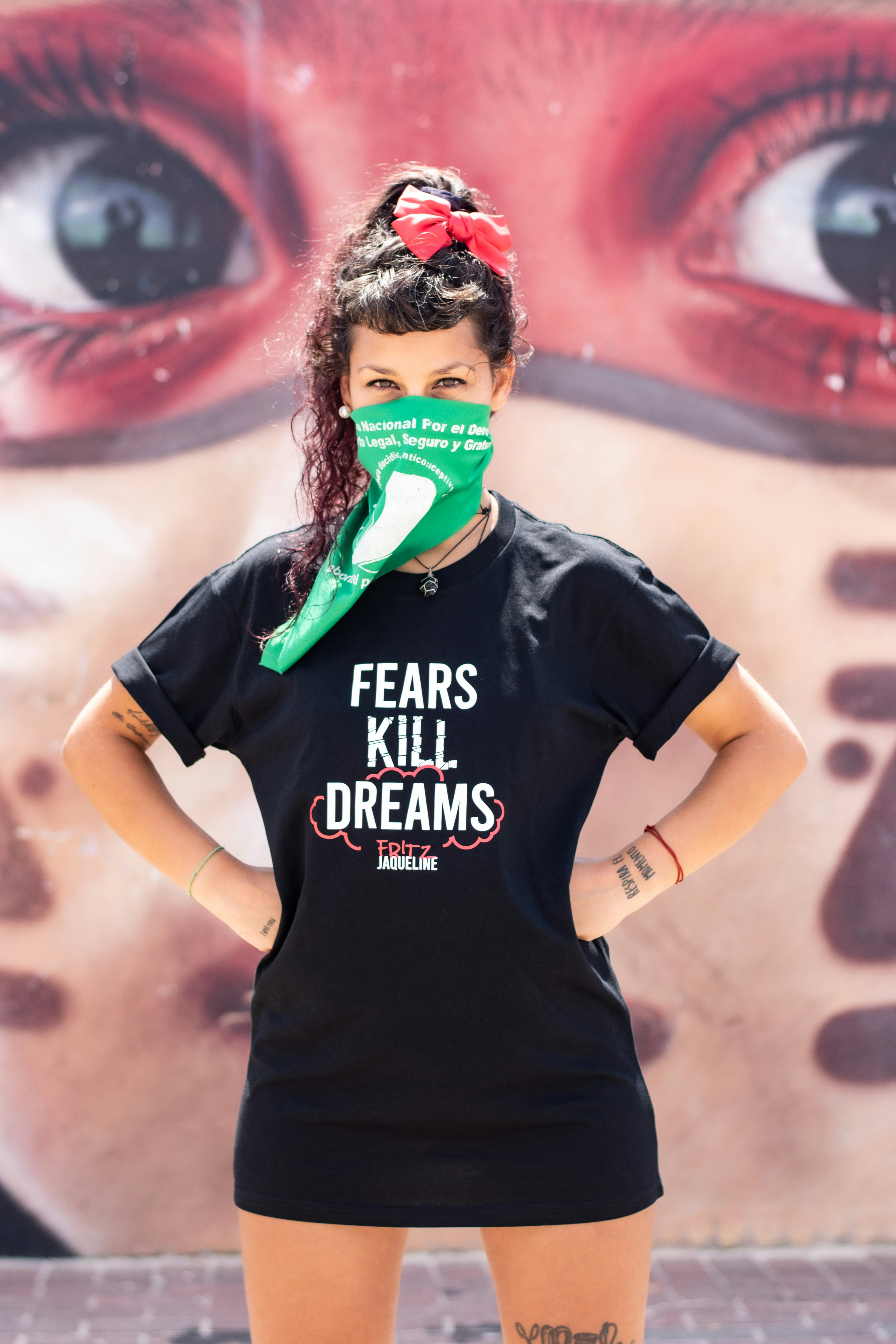 Bandana-clad protester stands before mural, defiant pose under large eyes.