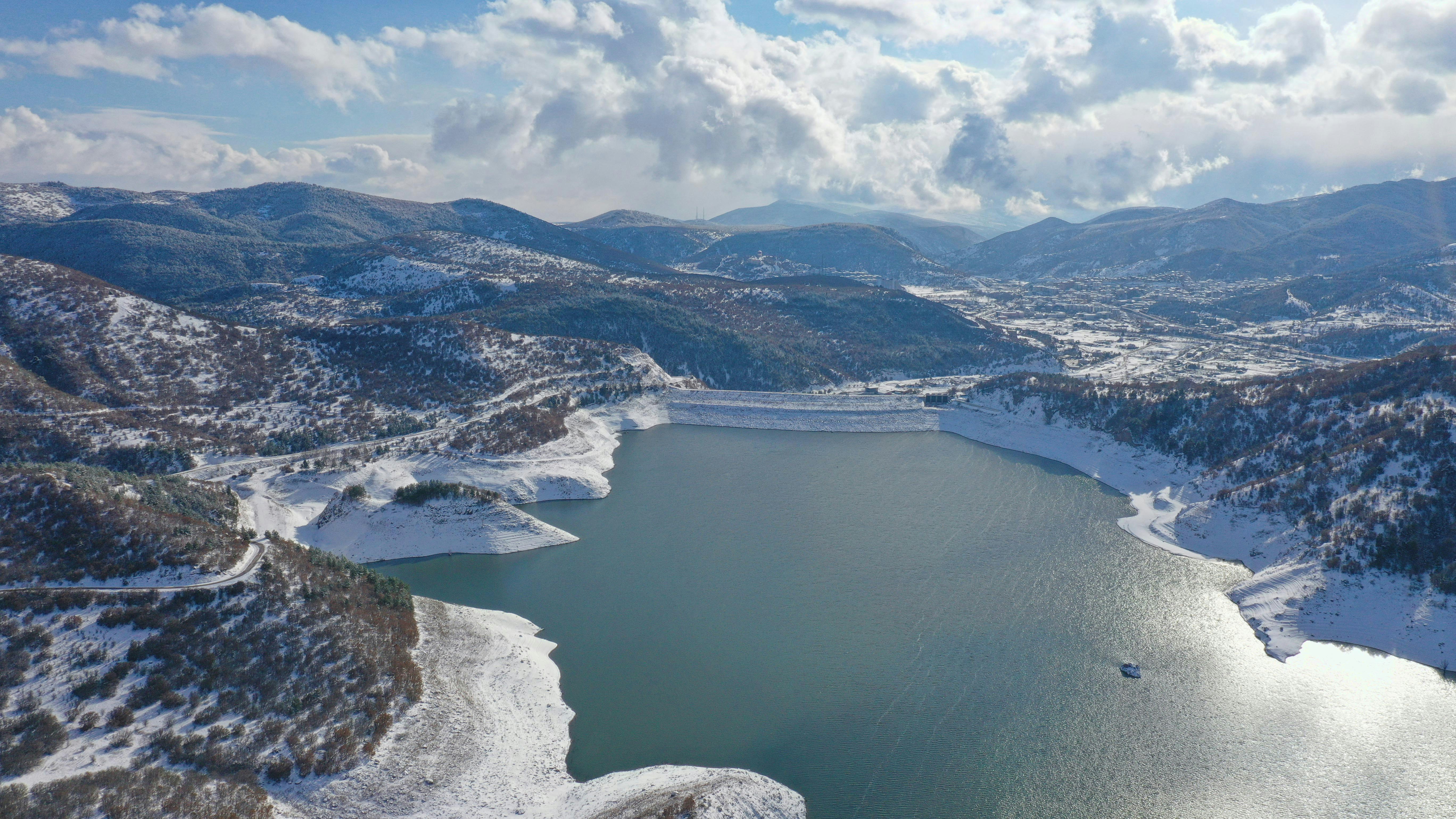 Snow-covered hills surround a partially frozen reservoir under a sky filled with scattered clouds.