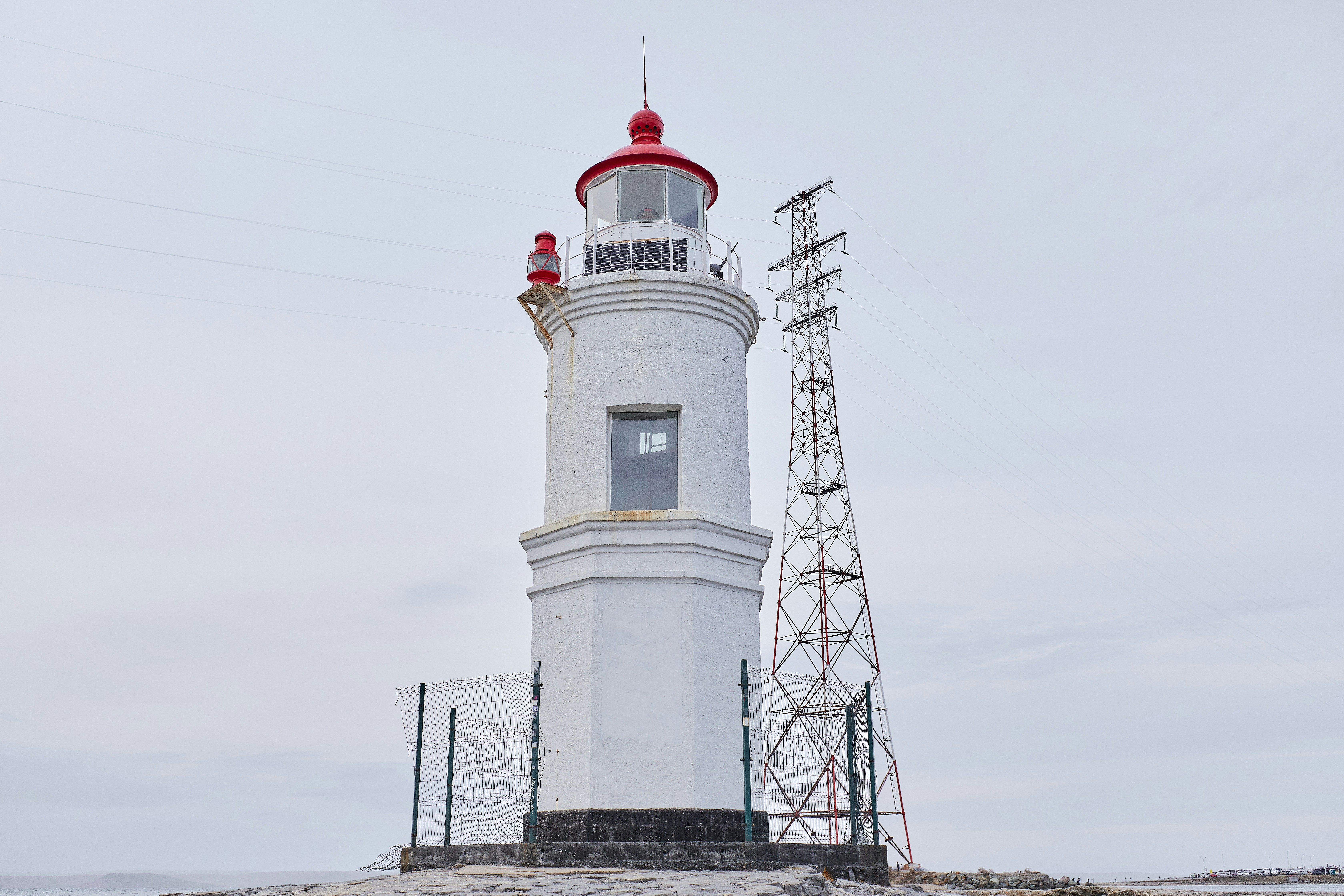 White lighthouse with red accents standing tall beside a communication tower against a muted sky.