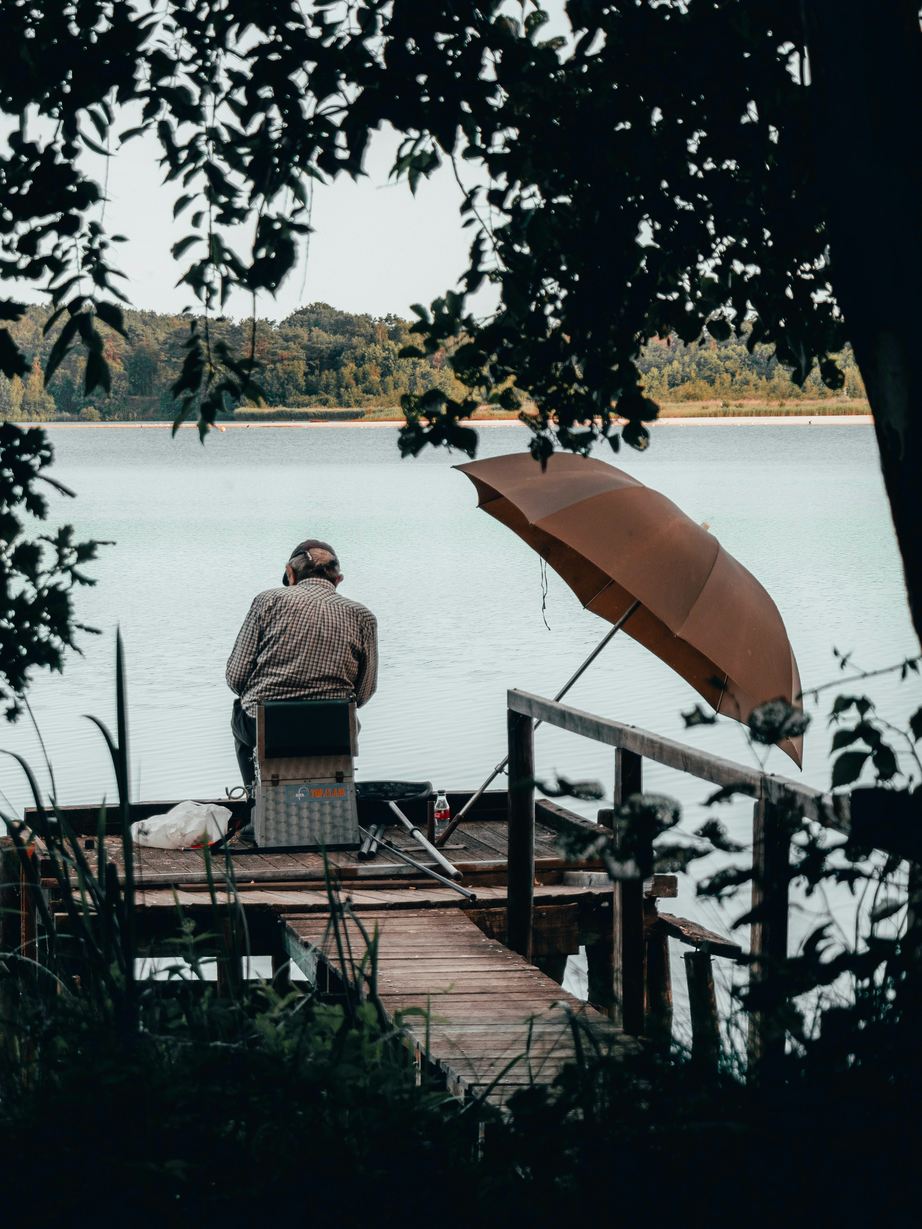 A solitary figure seated on a wooden dock, shaded by a large umbrella, gazes across a serene lake, surrounded by lush greenery.