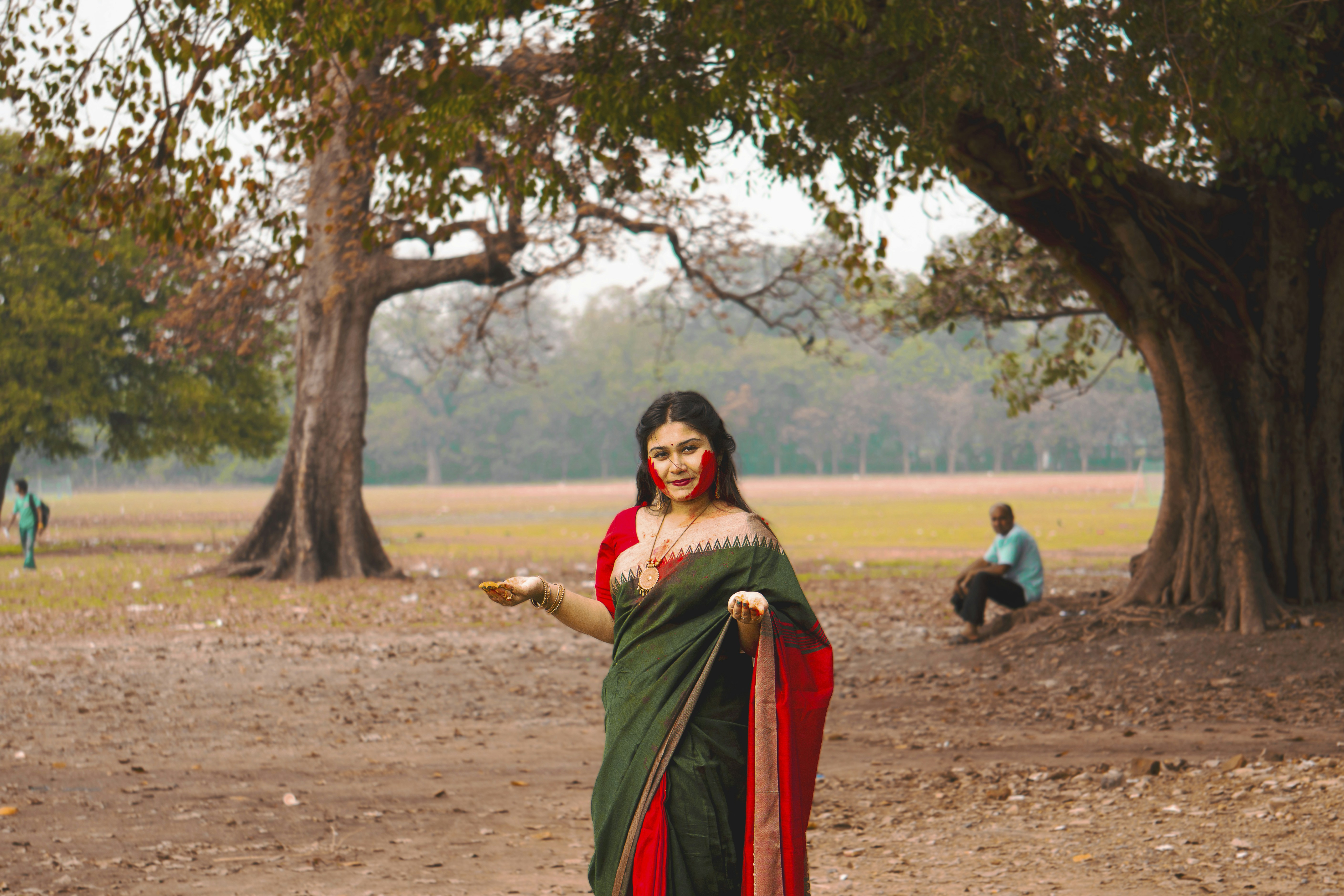 Woman in a green and red saree stands under sprawling trees at a park, holding colors in her hand.