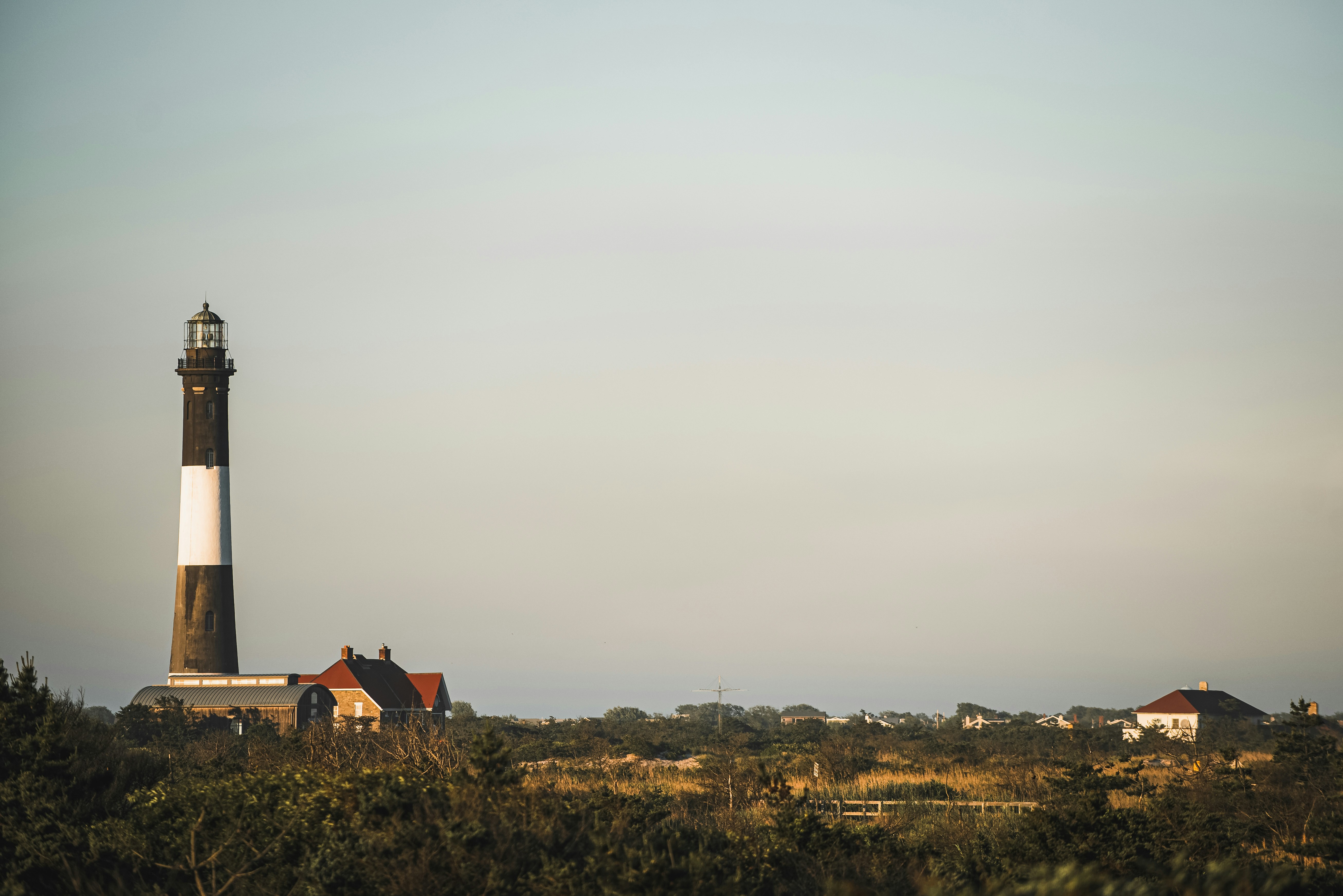 A historic lighthouse stands tall against a serene landscape, surrounded by gentle greenery and distant homes. The scene evokes a sense of calm and guidance.