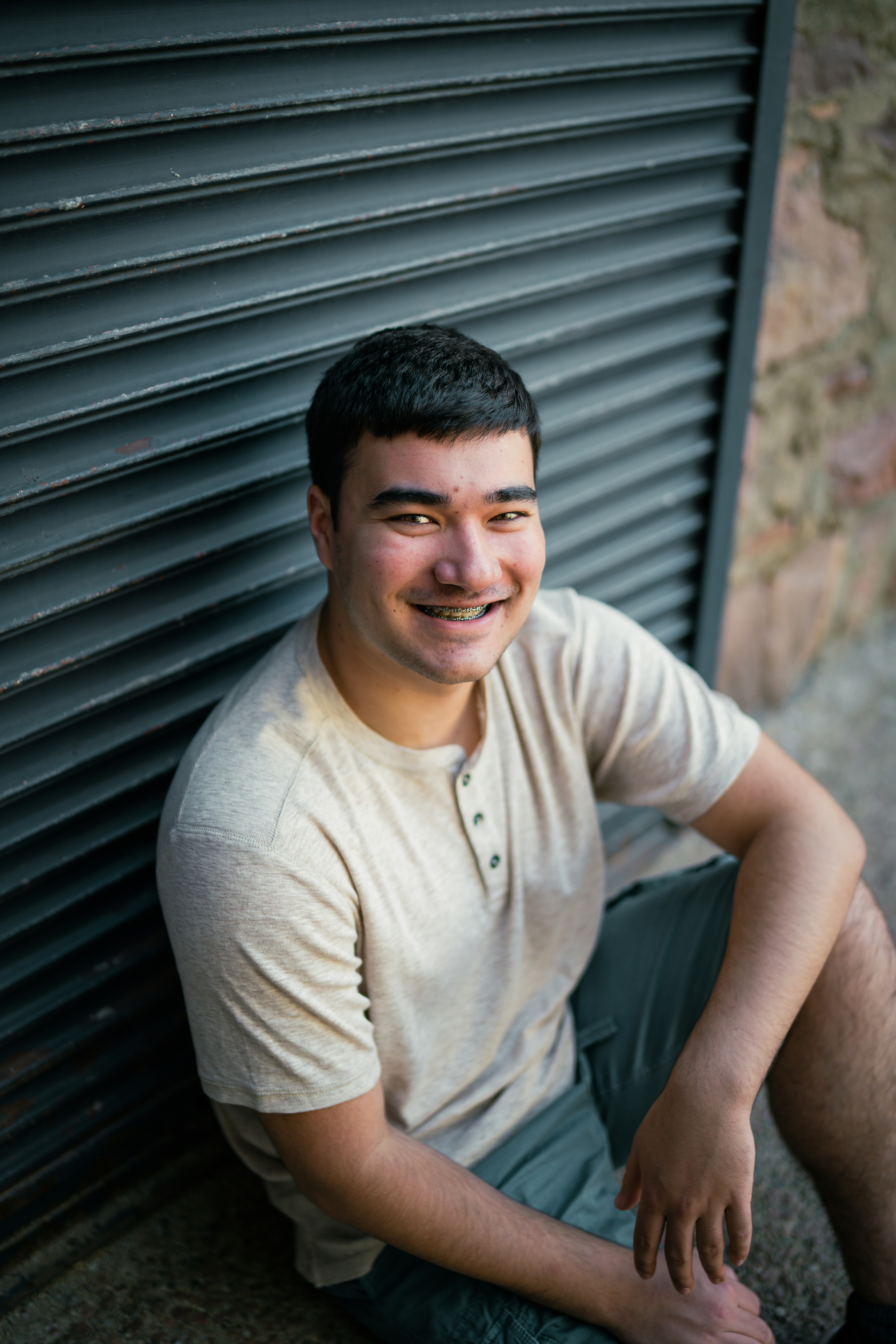 A young man smiles while seated against a textured wall with a shuttered background.