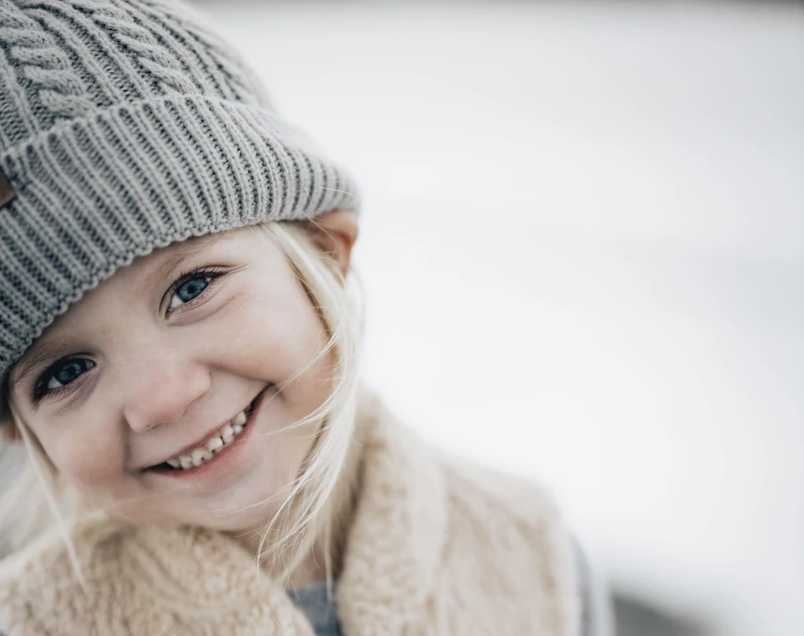 Young girl with a beaming smile, wearing a knitted hat and a cozy vest, set against a blurred winter backdrop.