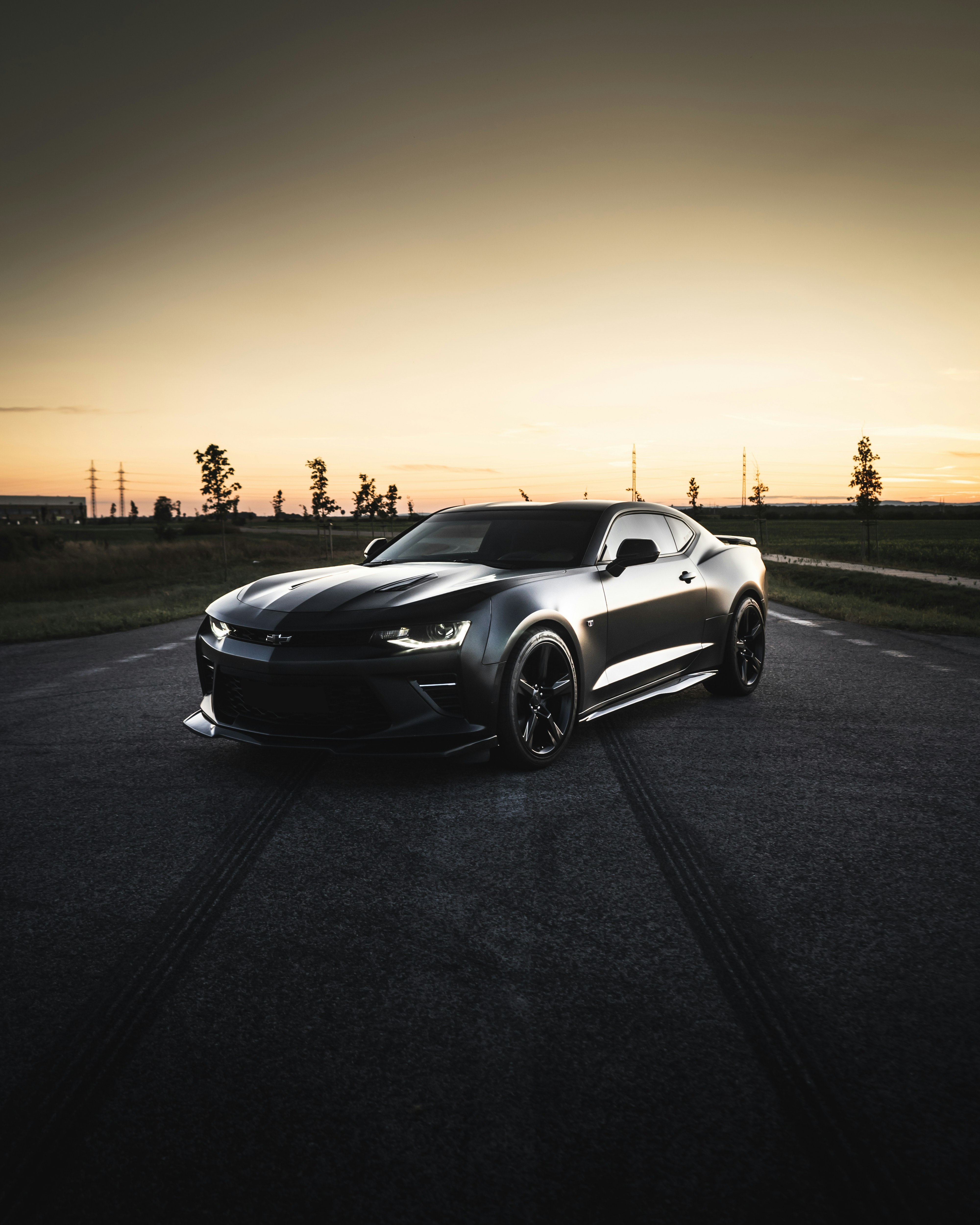 Sleek black sports car parked on an empty road at dusk, illuminated by a fading sunset. Tire tracks suggest recent speed and agility.