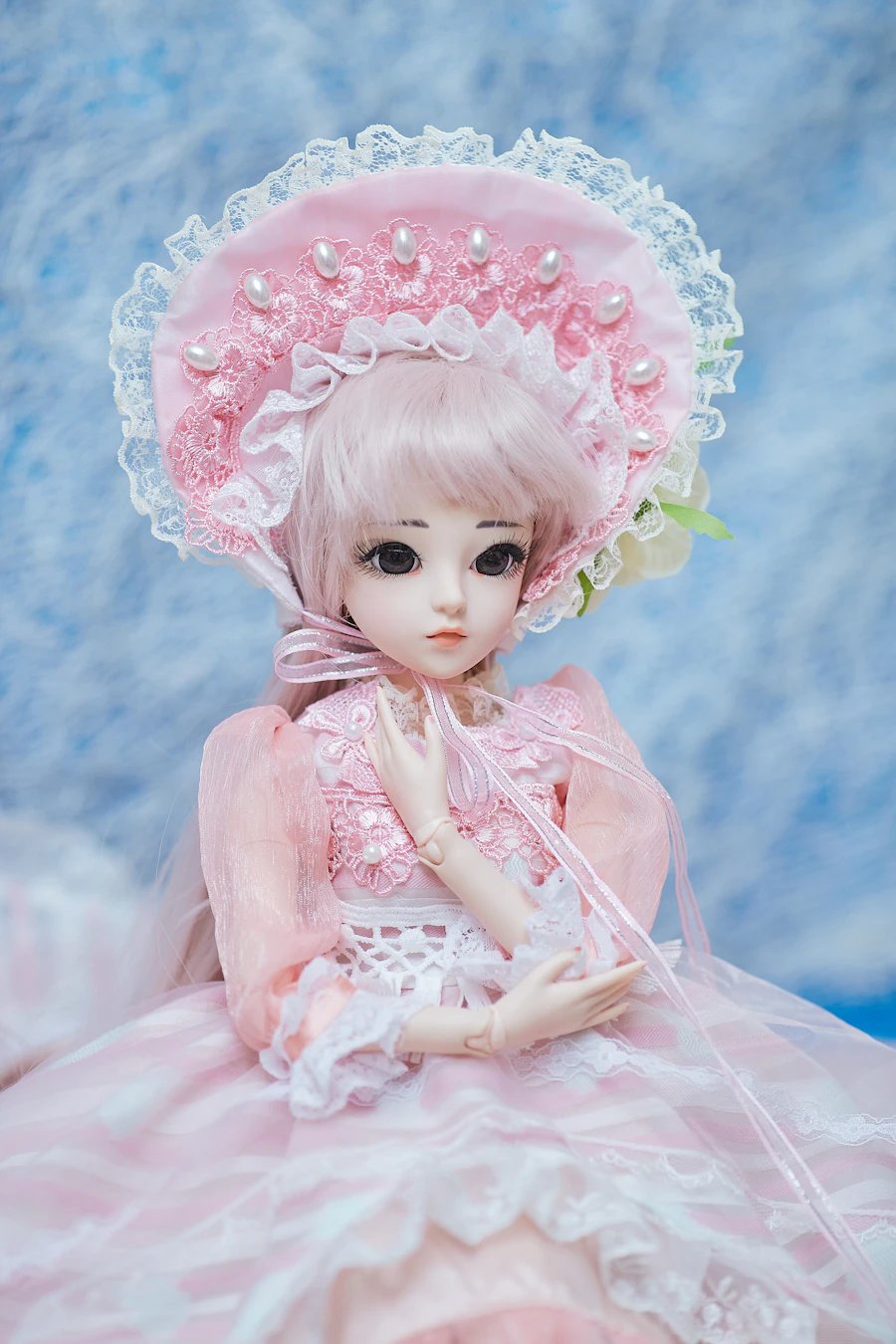 A porcelain-style doll with pink hair and a lace bonnet sits against a soft blue backdrop, dressed in a frilly pink dress with pearl accents.