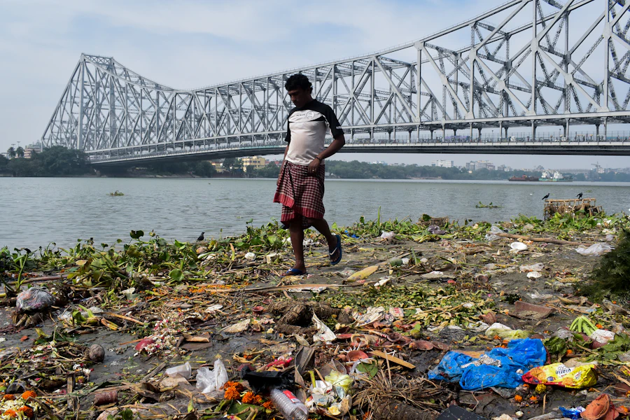 A person stands on a litter-covered riverbank in front of a large steel truss bridge spanning the water. The wide scene contrasts the industrial structure with the polluted foreground.