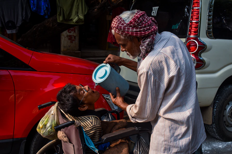 An older man in a turban pours water from a blue jug to a seated man in a wheelchair beside a red car. The scene captures a moment of care on a busy street.
