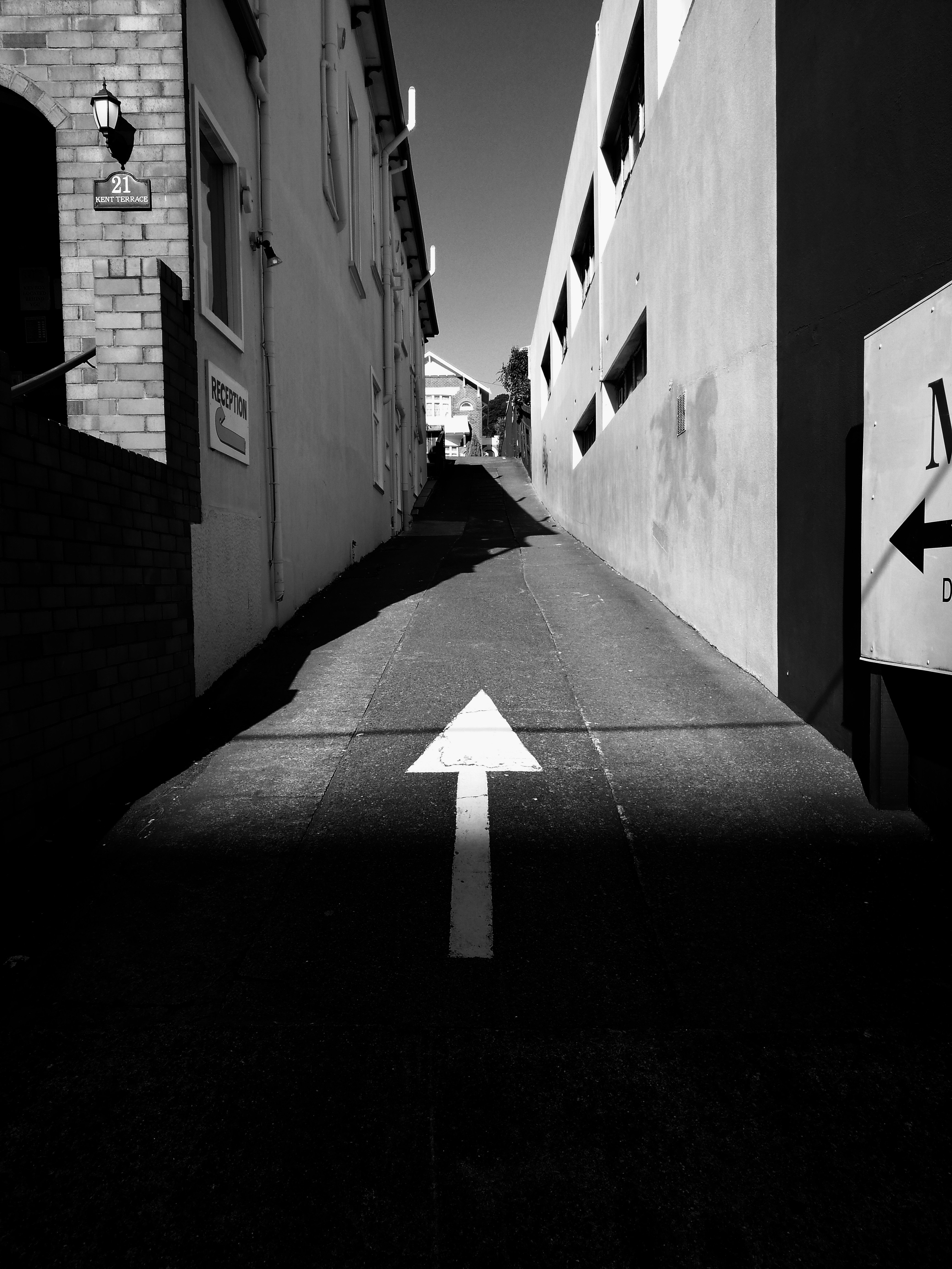 A stark black and white scene featuring a prominent arrow on a sloped pathway, flanked by buildings casting deep shadows. The composition invites exploration.