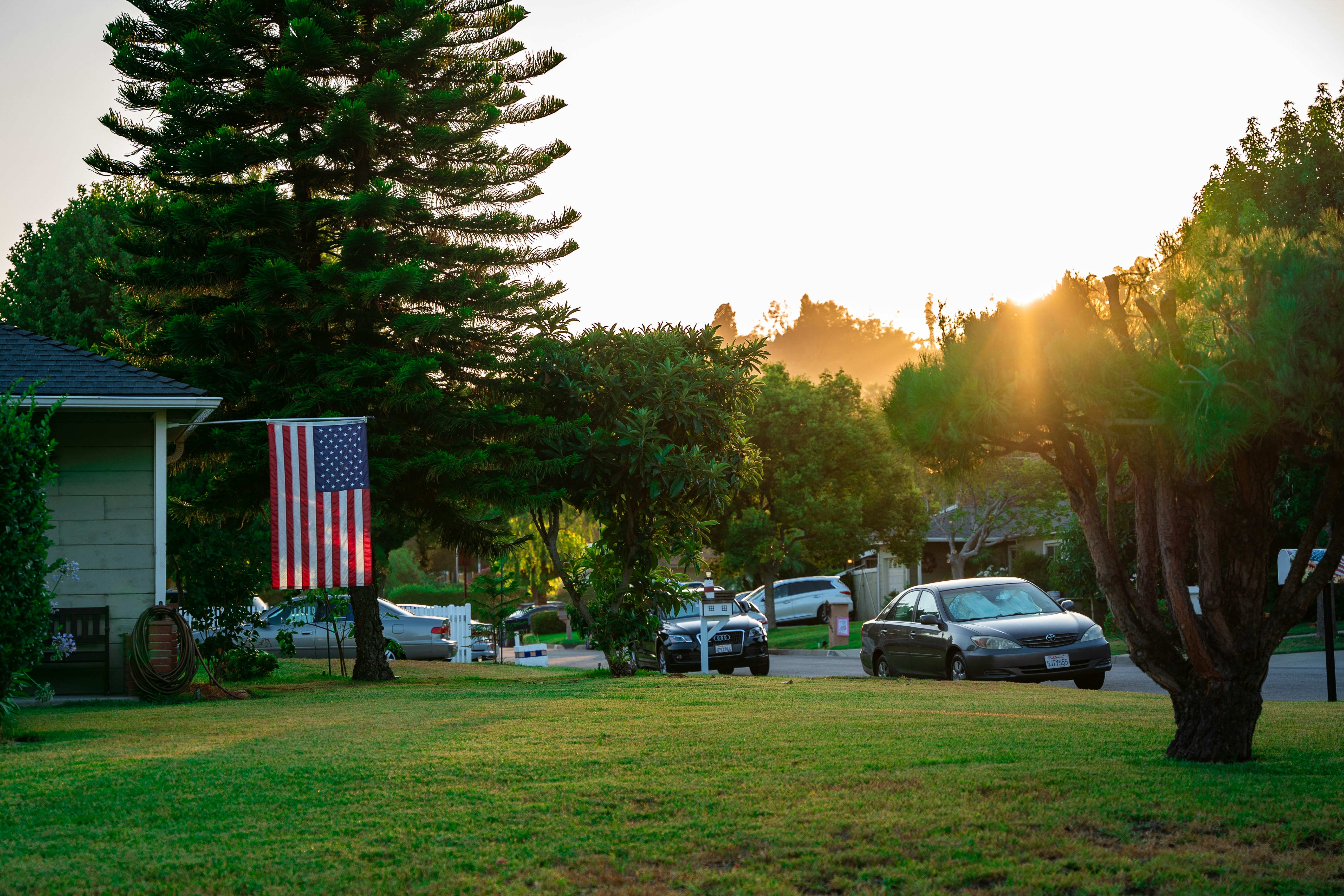 An American flag waves in the breeze, set against a backdrop of a sunlit neighborhood with cars parked along the street. The warm glow of sunset enhances the tranquil atmosphere.