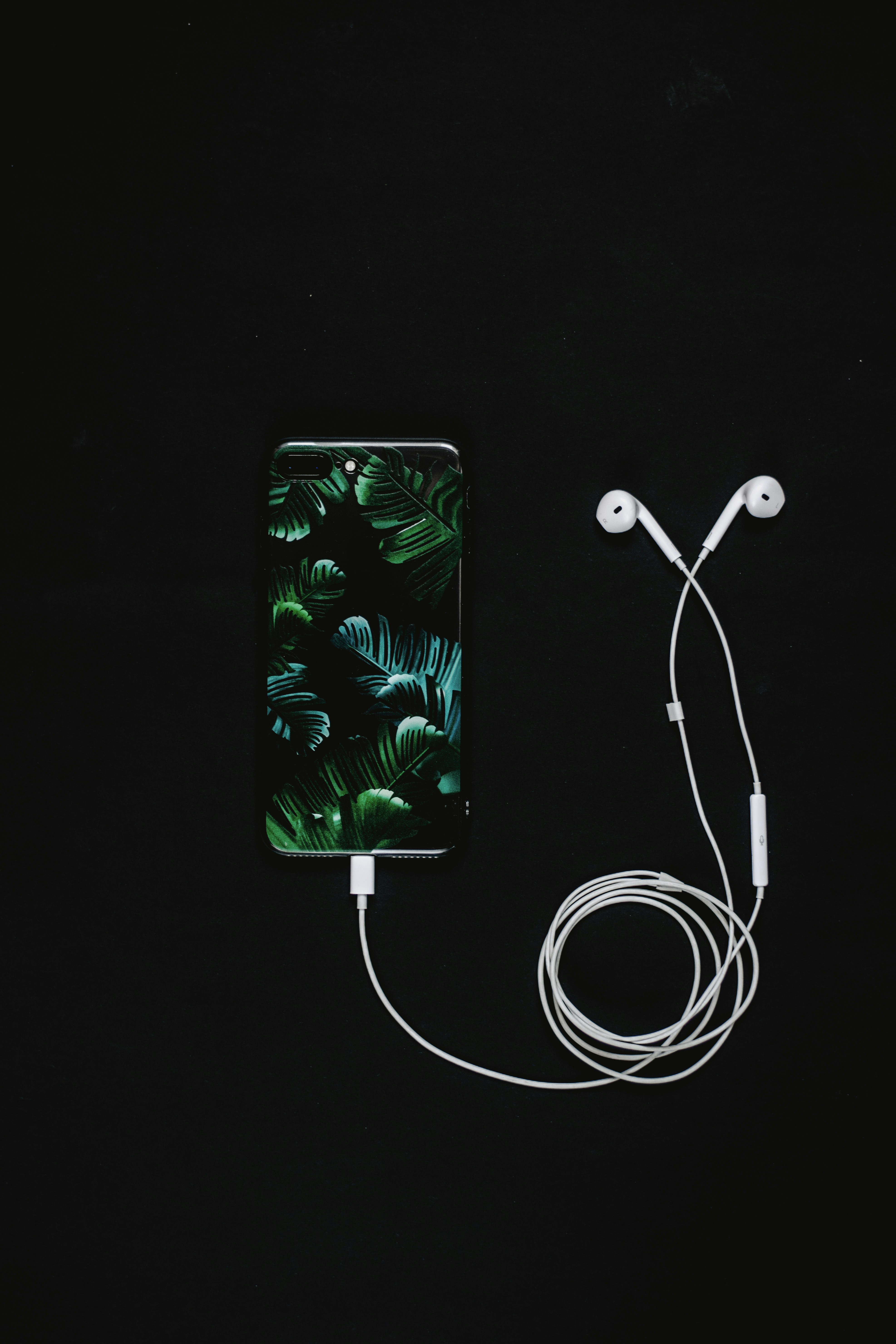 Smartphone with tropical leaf wallpaper connected to white wired earbuds rests on a dark surface. The setup emphasizes a clean, minimal composition with emphasis on texture and color.