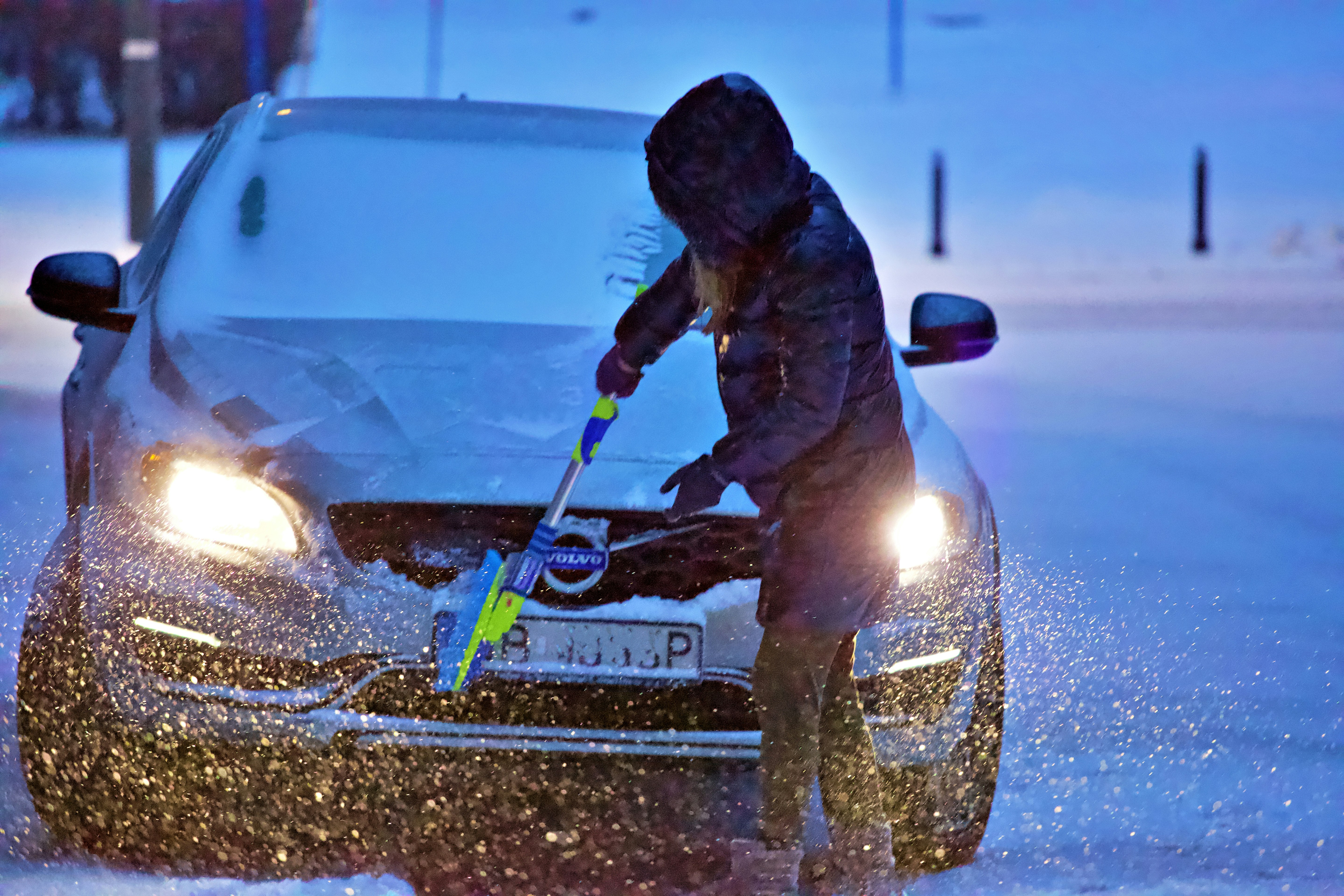 Person using a snow shovel to clear snow from a car parked in a snowy street during twilight.