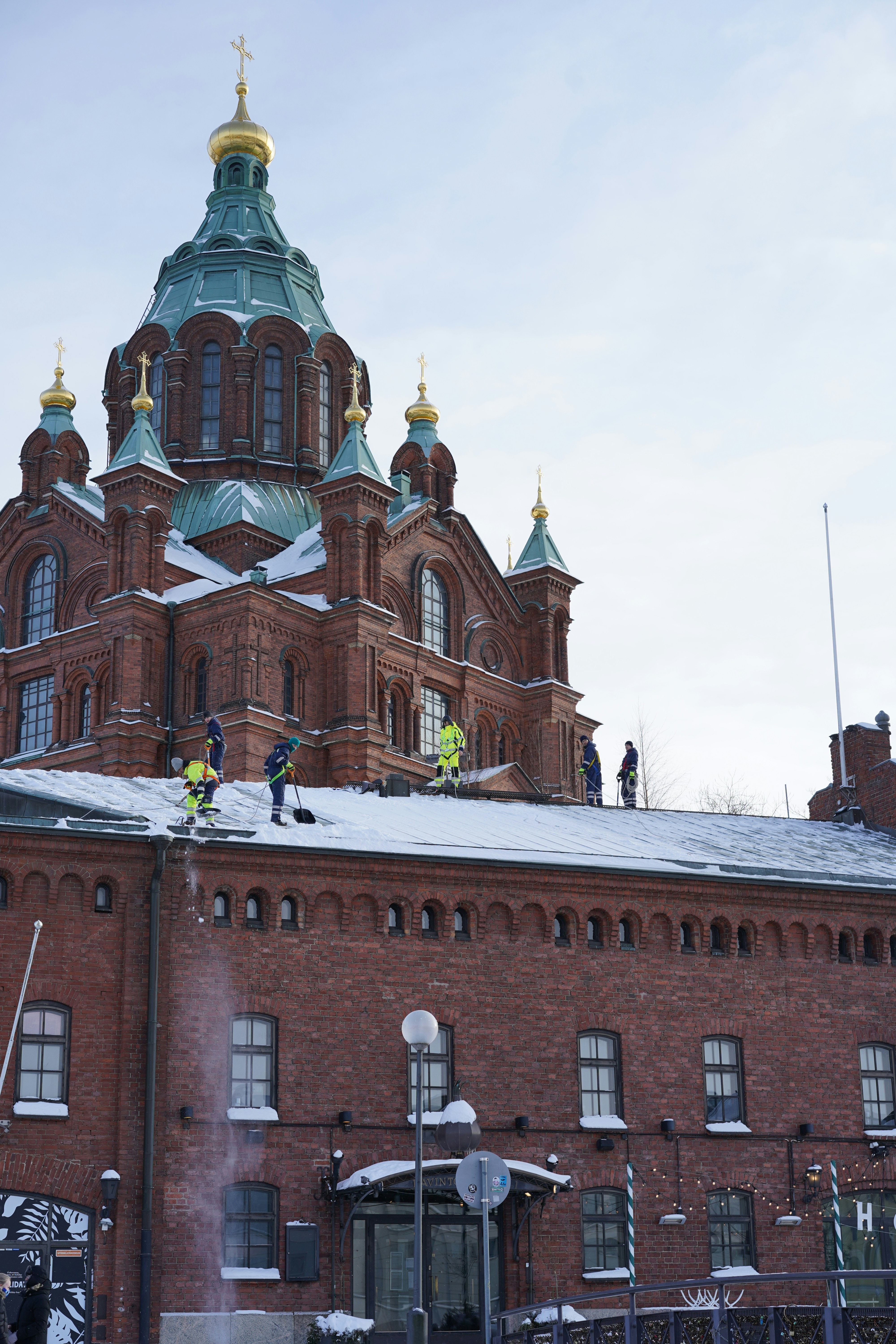 Workers in bright gear removing snow from a historic building's roof, highlighting the challenges of winter maintenance.