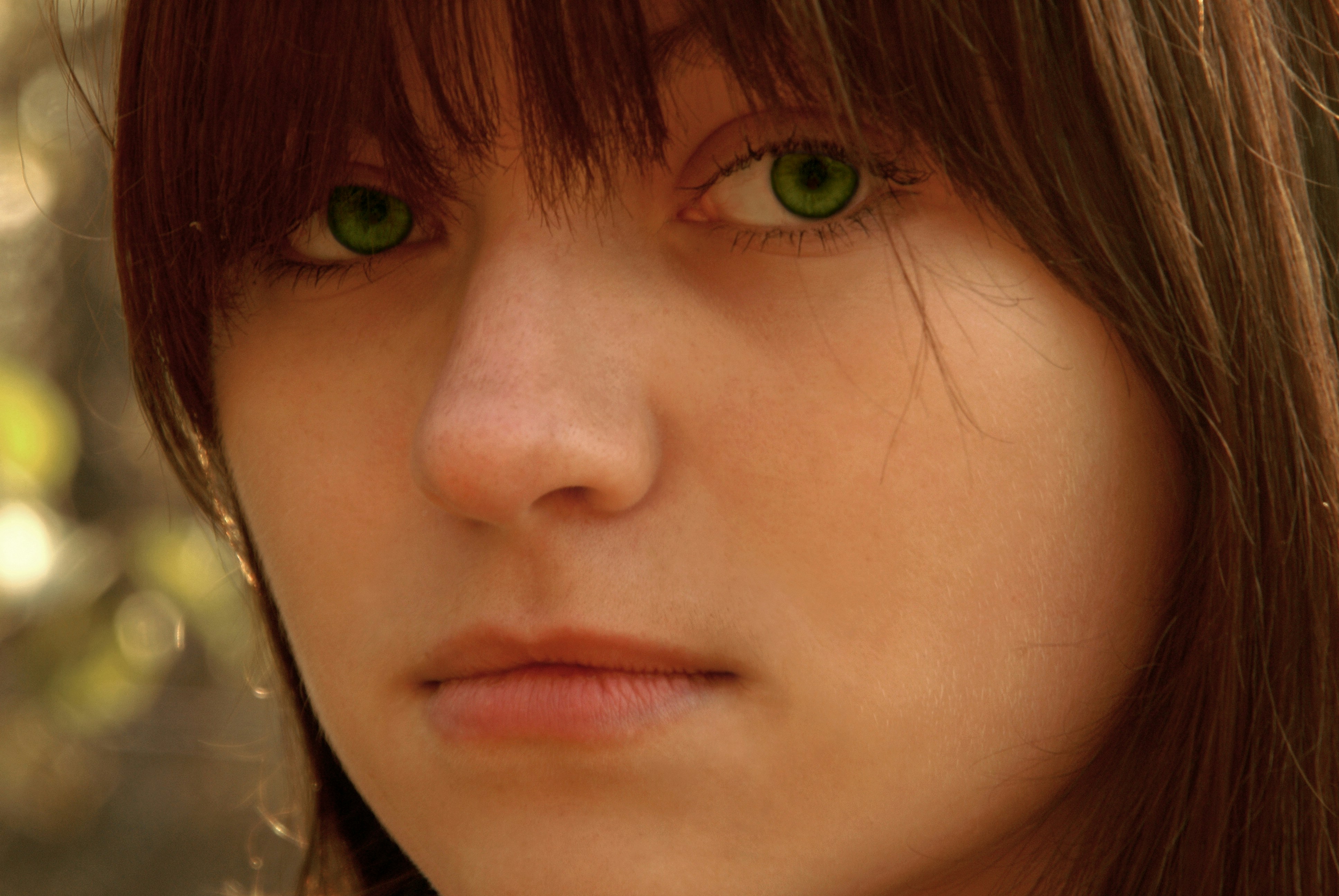 Close-up portrait of a young woman with striking green eyes and dark hair, set against a softly blurred natural background.
