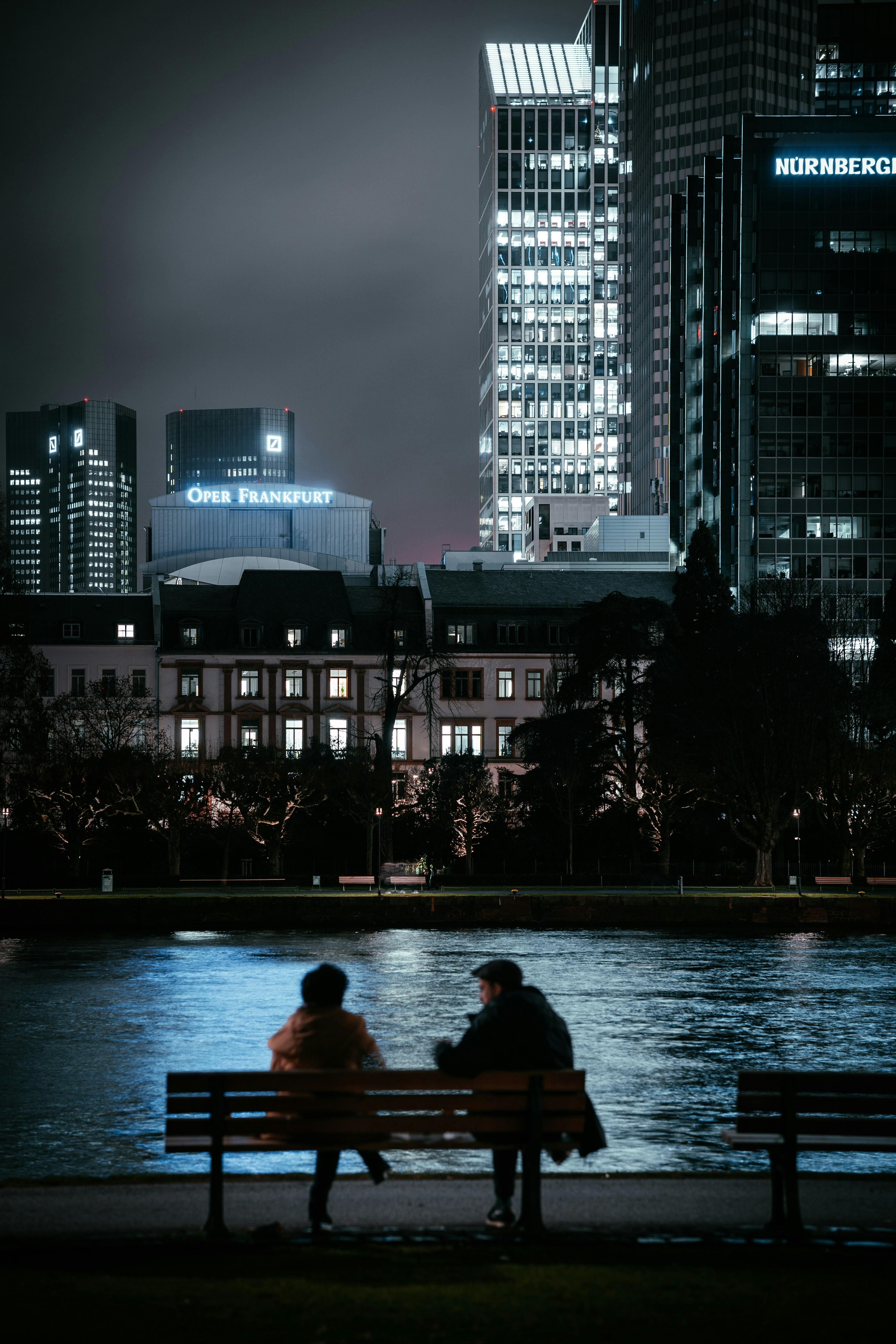 Two figures seated on a bench along the riverbank, illuminated by city lights reflecting on the water. The backdrop features modern skyscrapers and historic architecture.