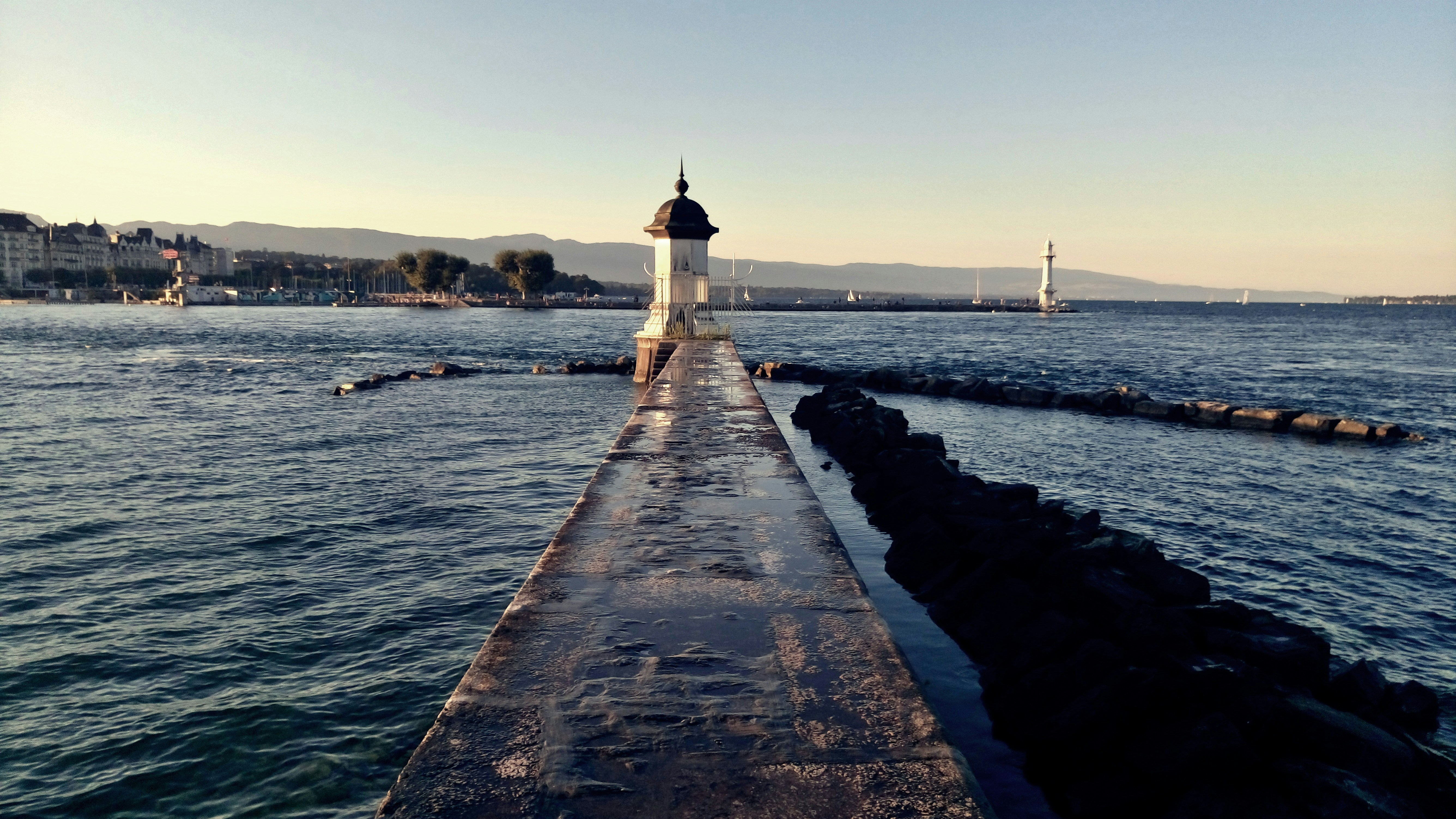 Narrow stone walkway leading to a lighthouse with calm waters and distant mountains at dusk.