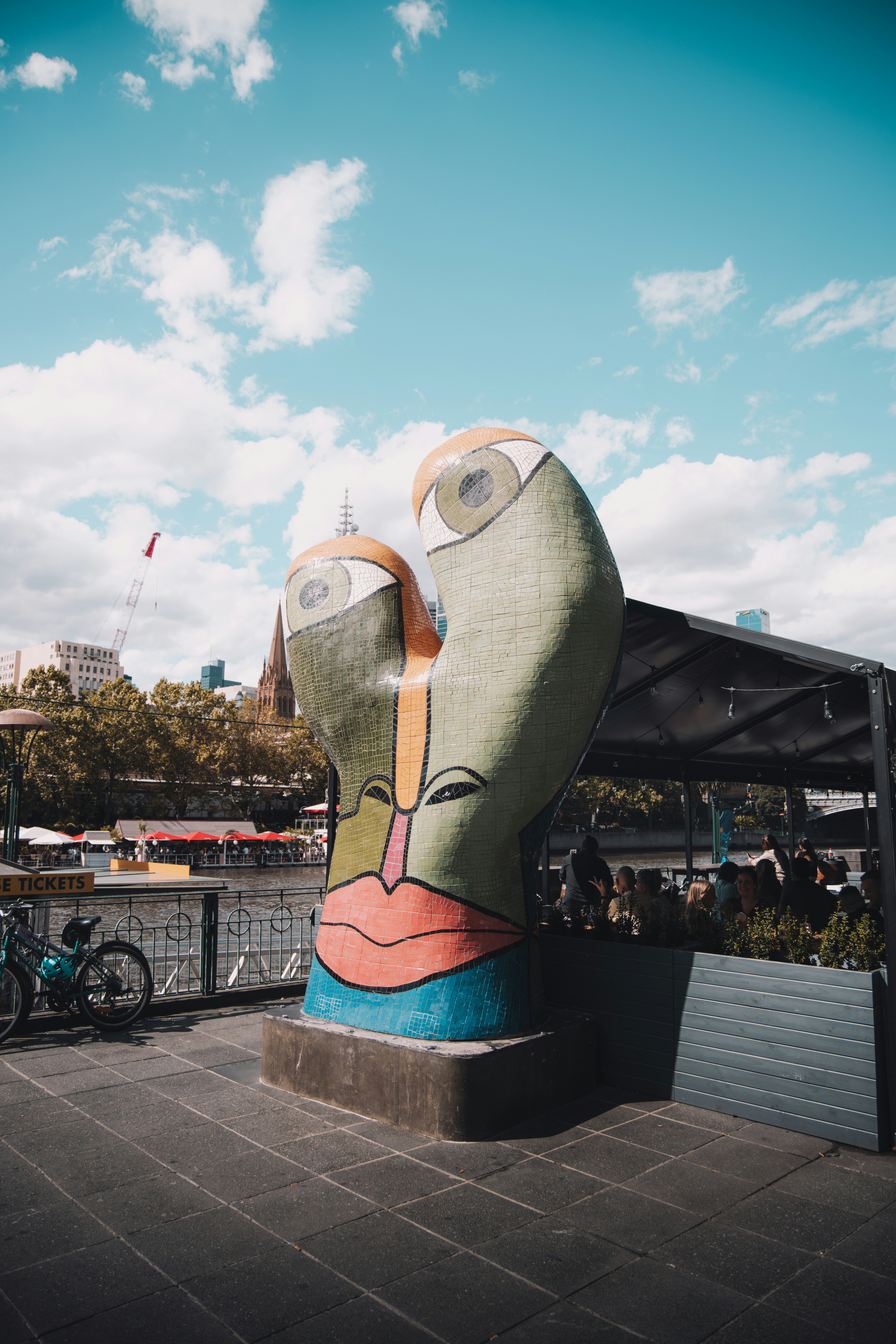 Vibrant sculpture with exaggerated facial features stands prominently near a riverside café, inviting curiosity and interaction.