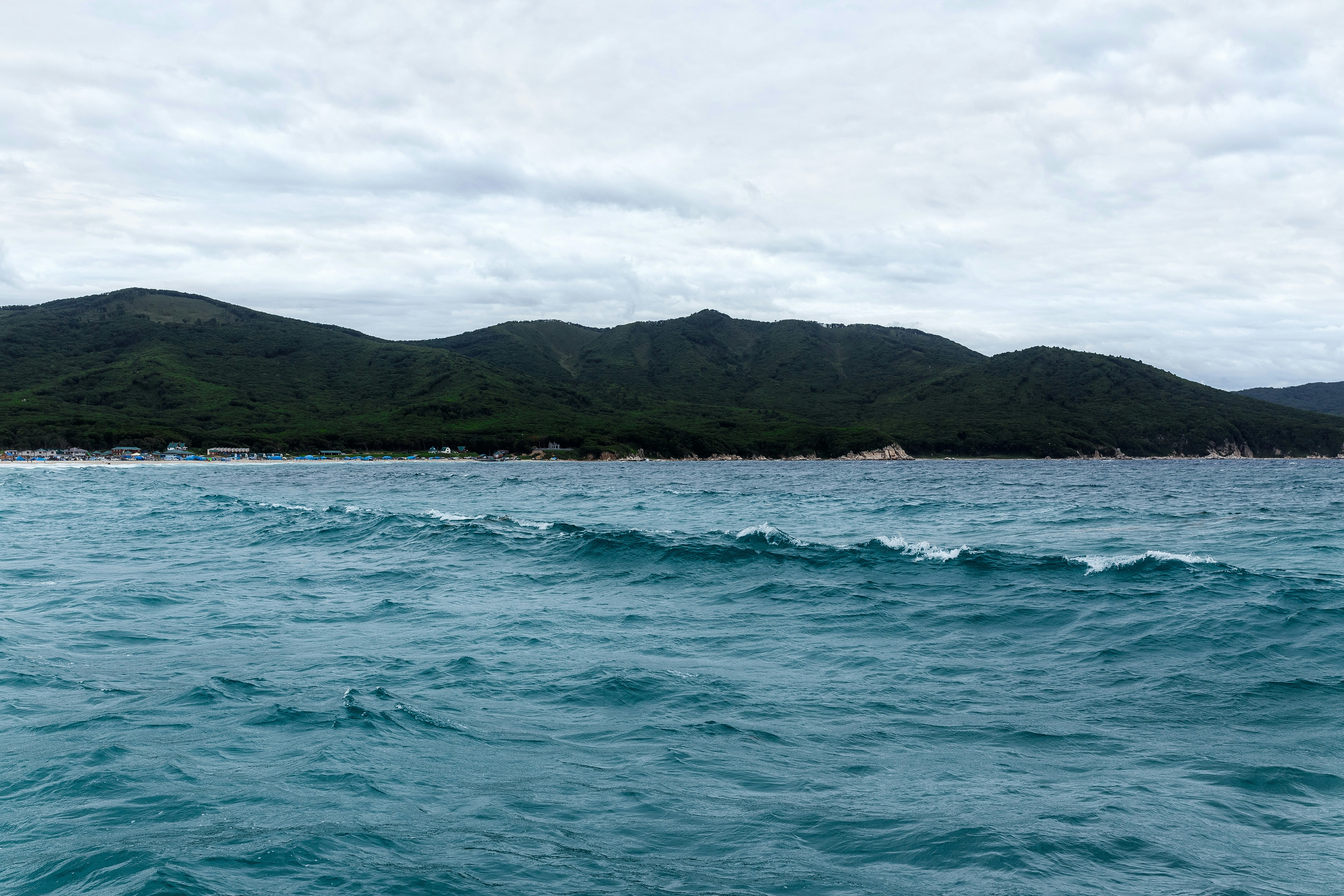 Waves gently lap against the shore, framed by lush green mountains under a cloudy sky.