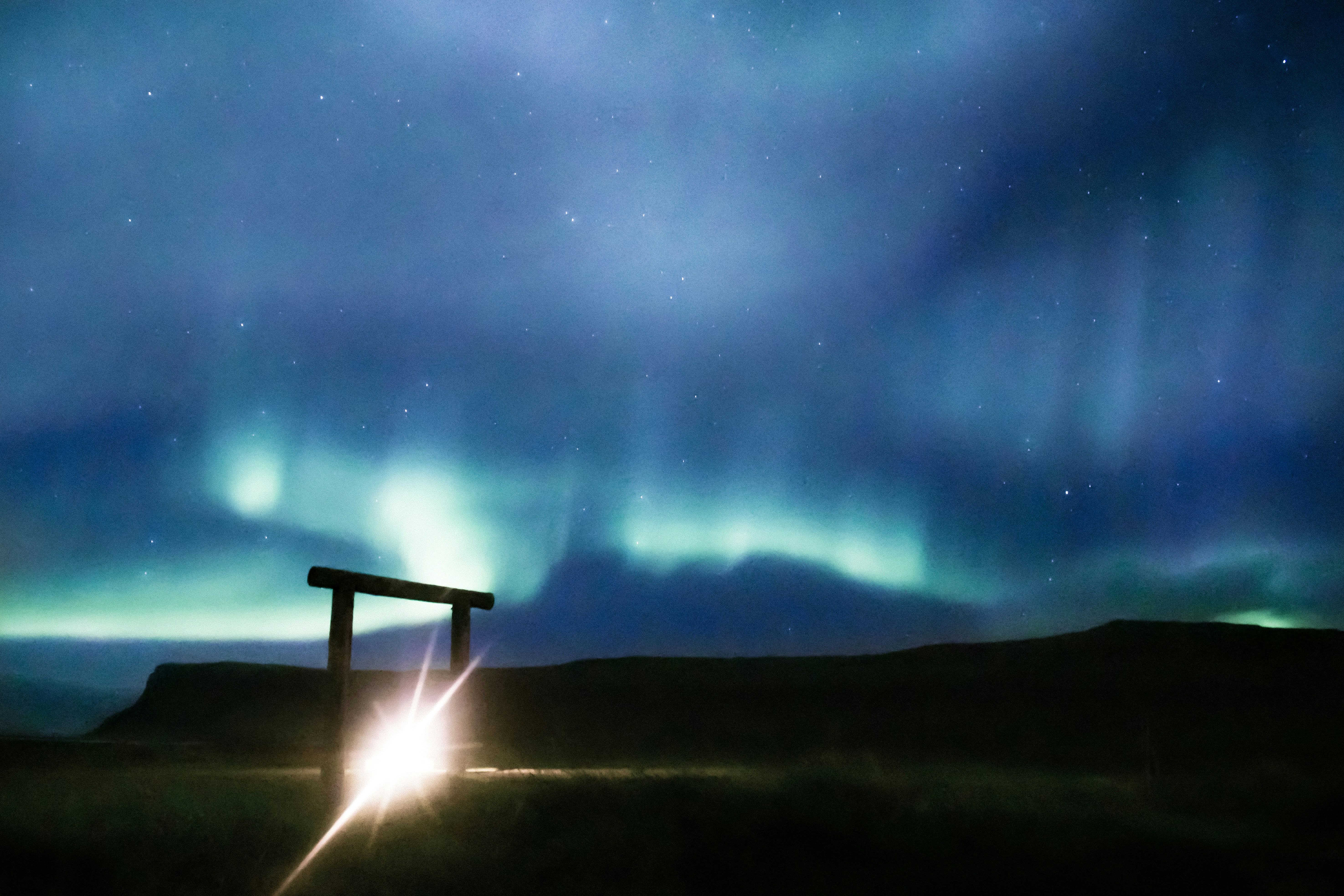 A torii gate illuminated by a bright light under a mesmerizing display of the aurora borealis, casting an ethereal glow against the night sky.