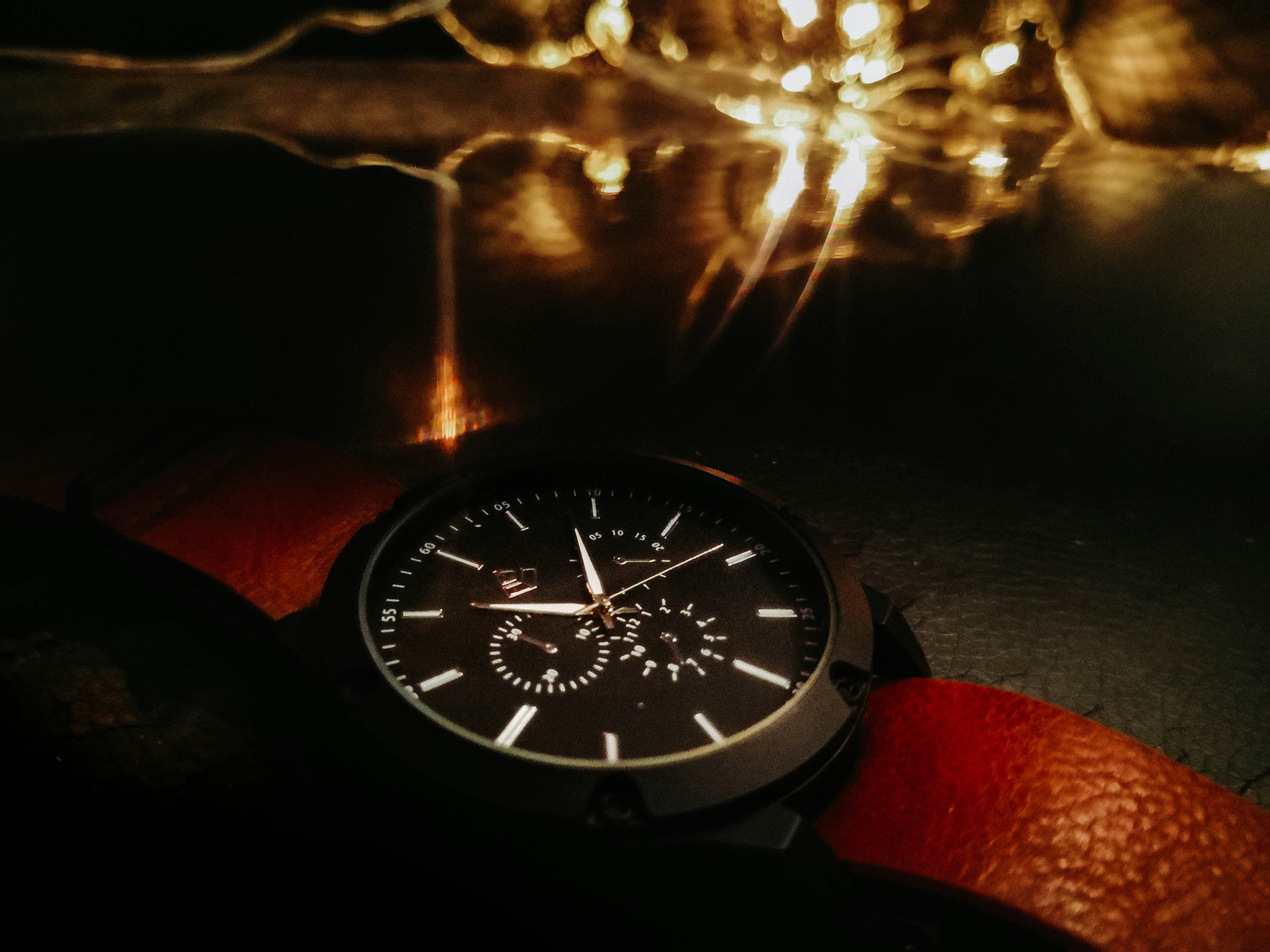 Close-up of a black chronograph watch resting on a leather strap, illuminated by soft, warm fairy lights in the background.