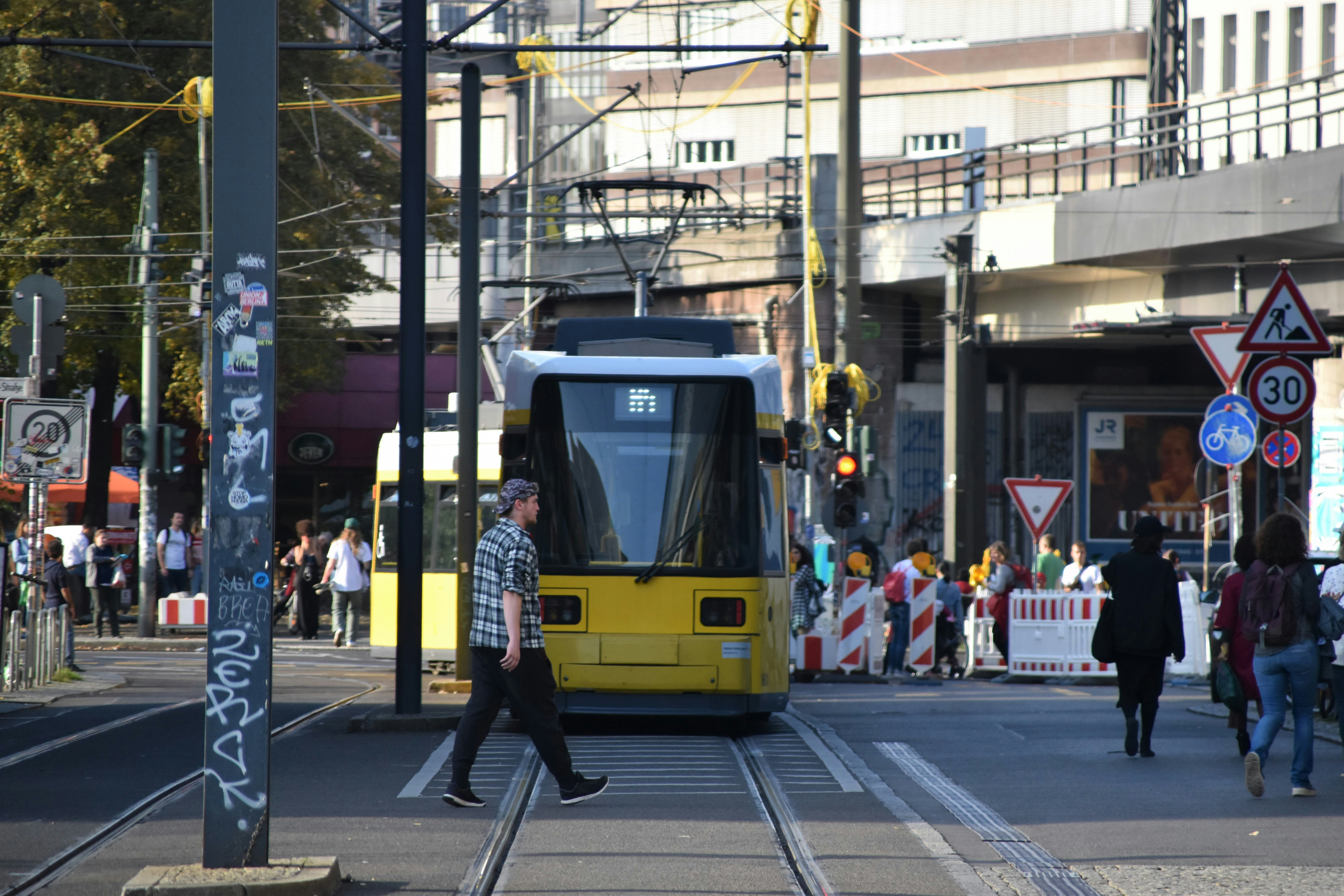 Tram approaching a busy intersection with pedestrians navigating the urban landscape. Traffic signs and construction barriers reflect the dynamic city environment.