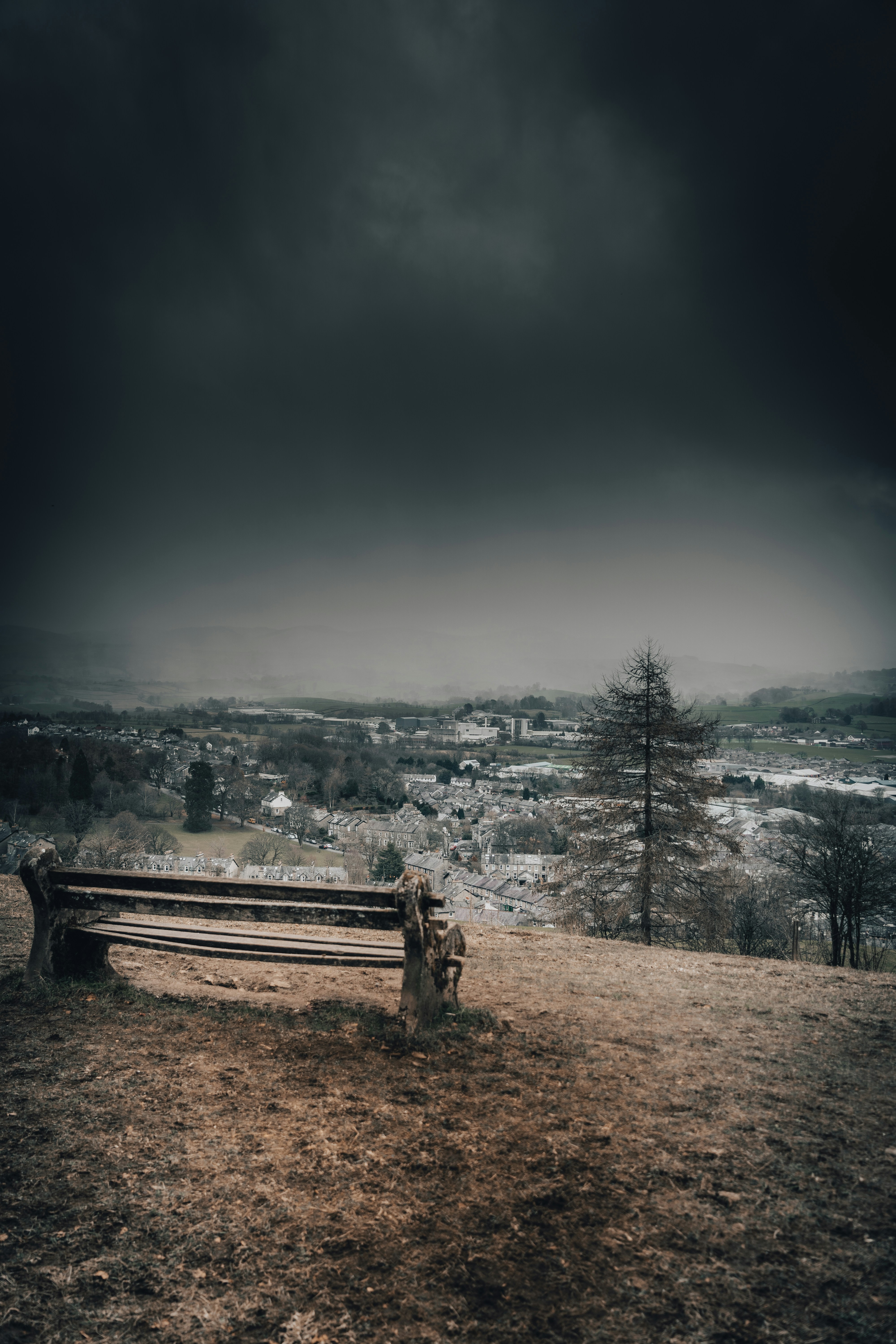 Weathered wooden bench overlooking a tranquil valley under a moody sky. The distant landscape reveals a small town nestled among the hills.