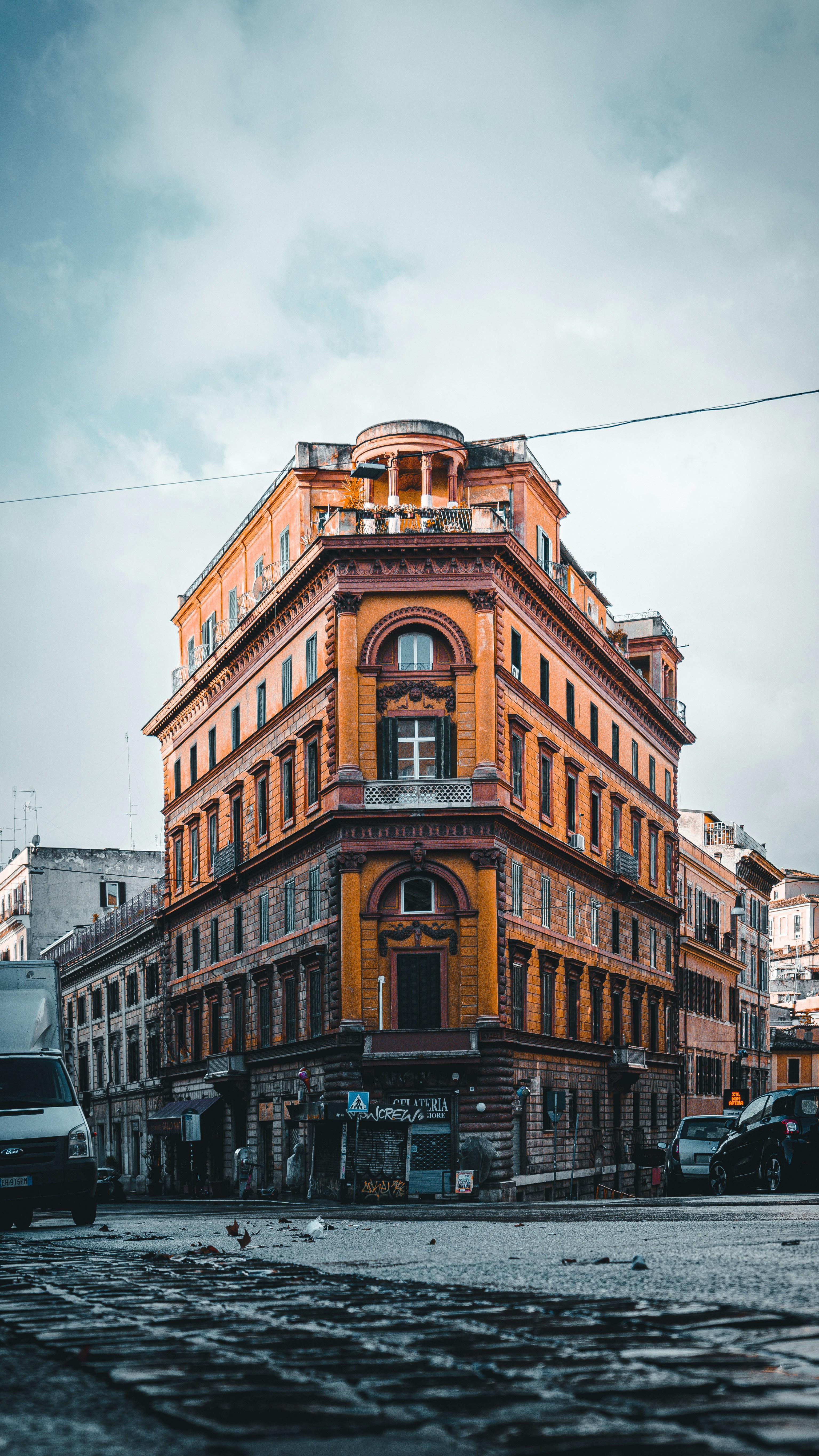 Historic orange building with ornate architectural details stands prominently at a city intersection, framed by a cloudy sky.