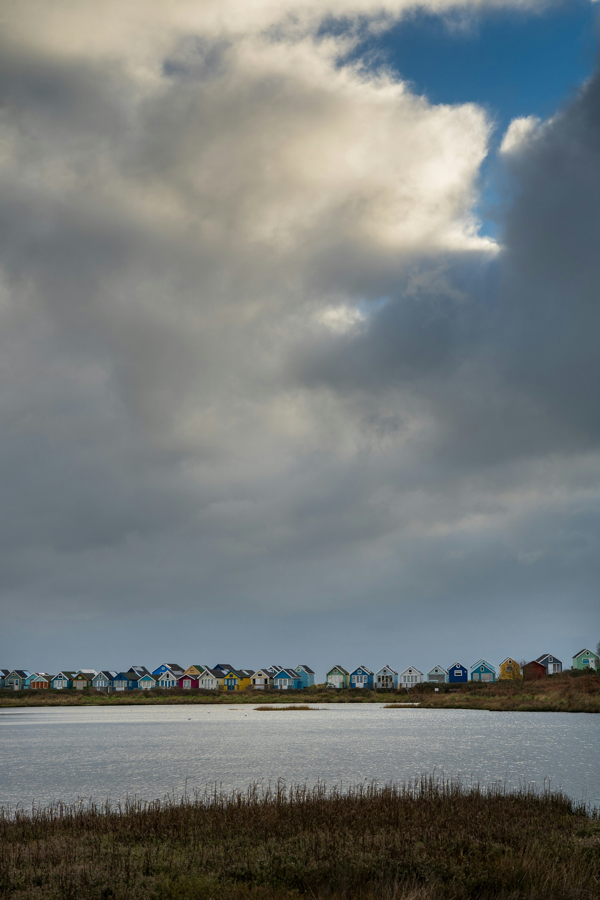 Colorful beach huts line the shore beneath a dramatic cloudy sky at Mudeford Spit, Dorset.