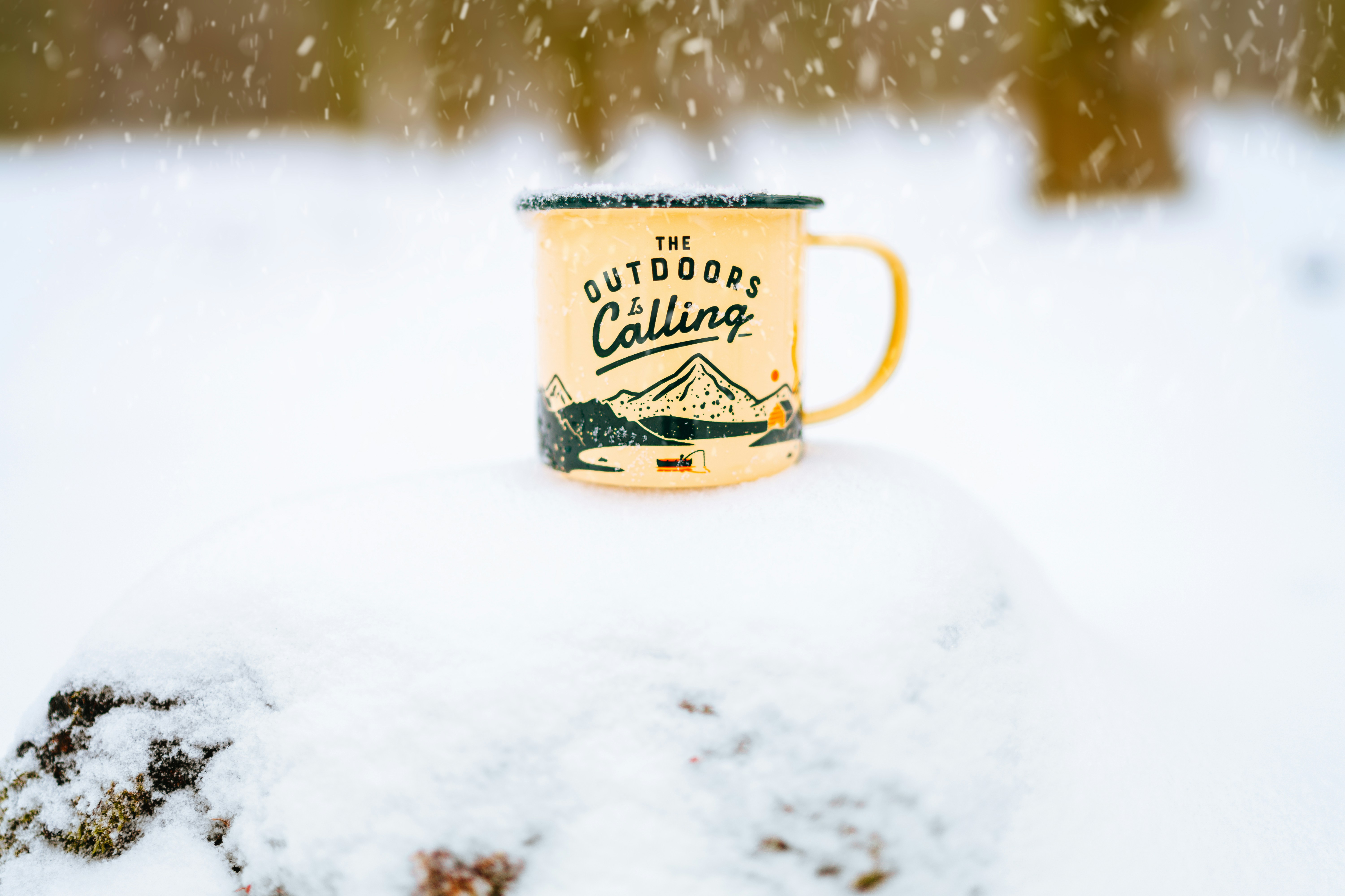 A vintage-style camping mug rests atop a snow-covered rock, surrounded by a gentle snowfall, evoking a sense of winter exploration.