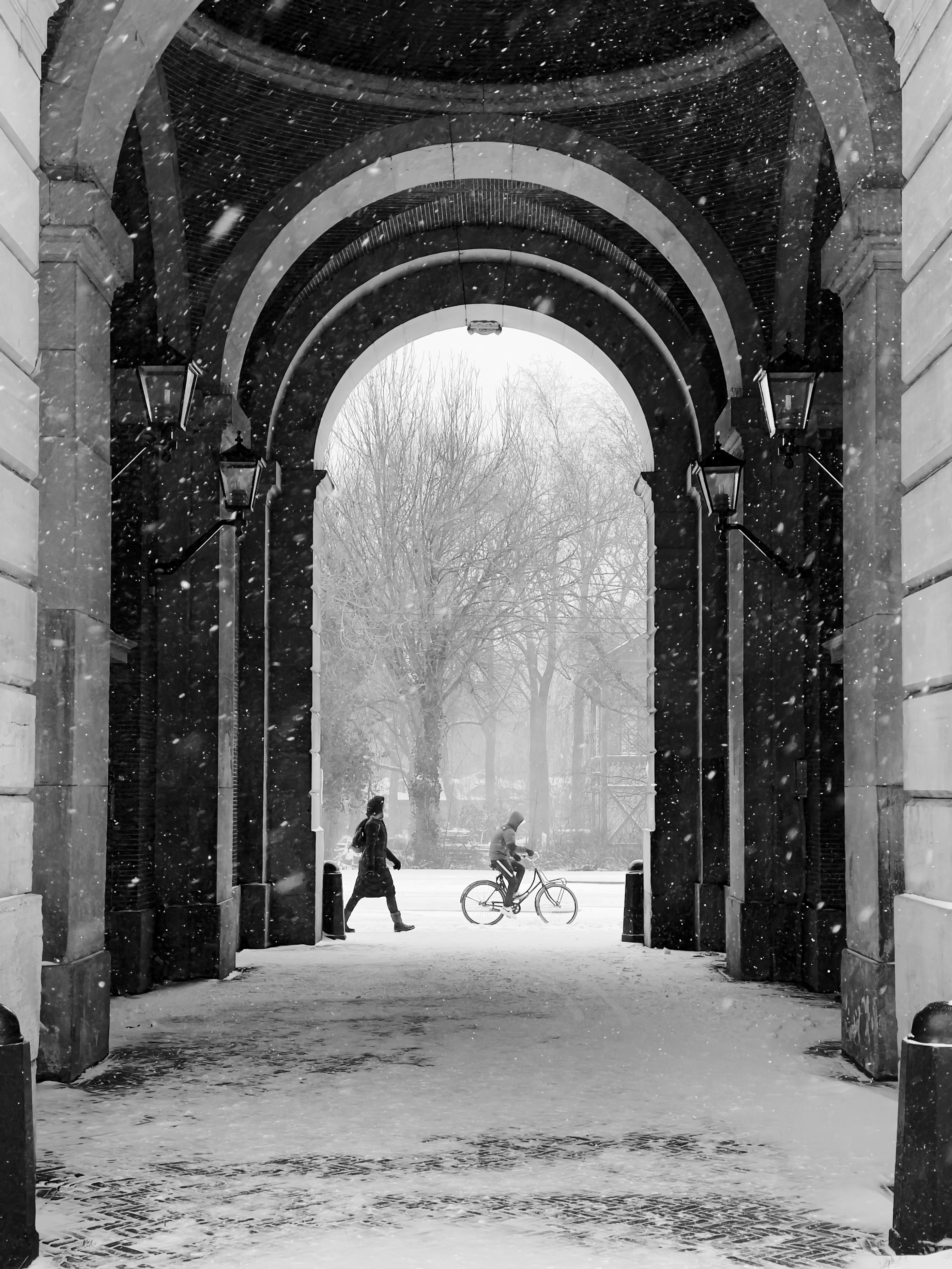A pedestrian and a cyclist navigate through a snowy archway, surrounded by softly falling snowflakes. The scene captures the essence of winter in a monochromatic palette.
