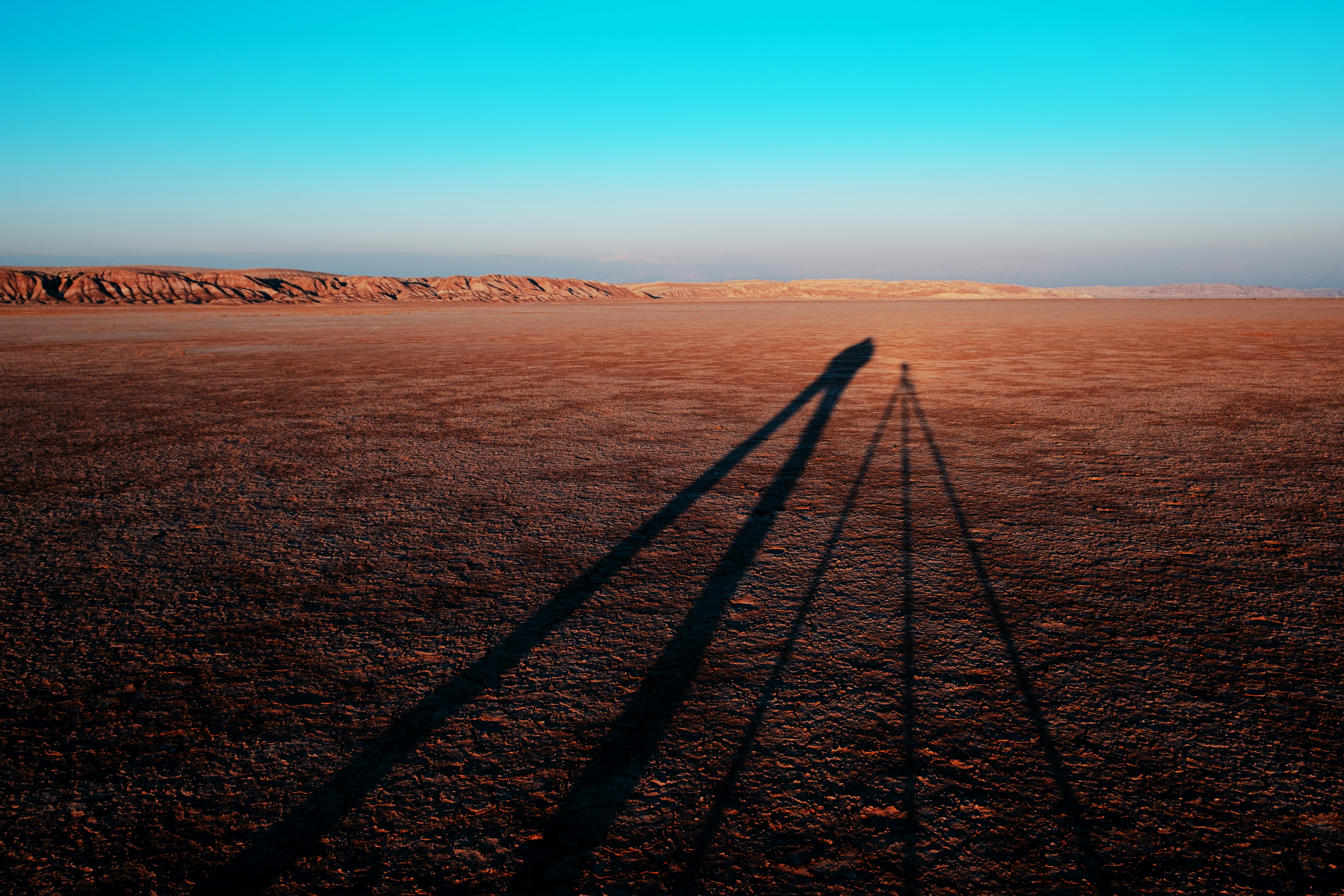 Long shadows stretch across a vast salt flat under a clear blue sky, creating a stark contrast between the earth and the horizon.