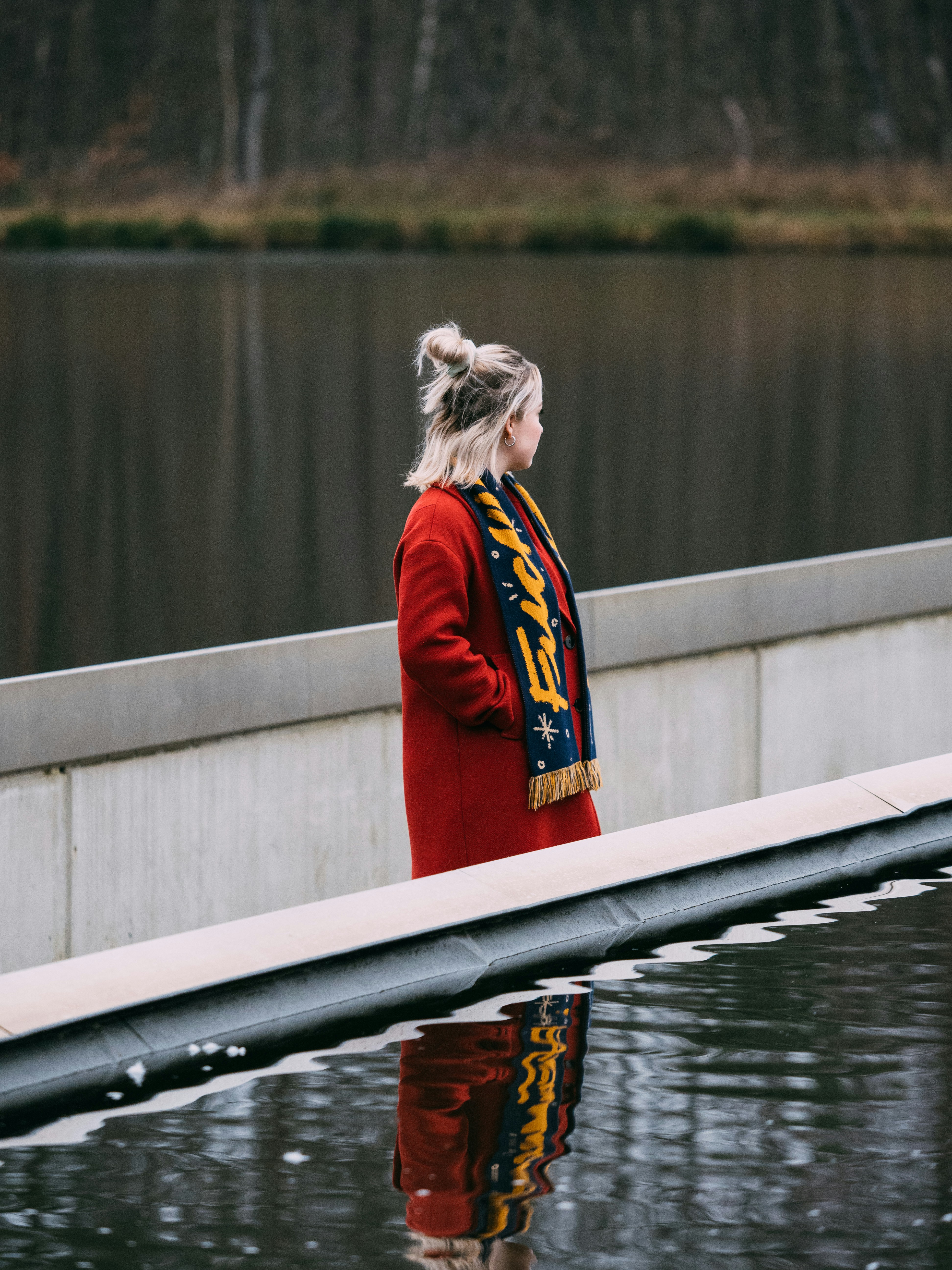 A woman in a vibrant red coat stands by a calm body of water, her reflection mirrored in the stillness. The scene captures a moment of contemplation amidst nature.